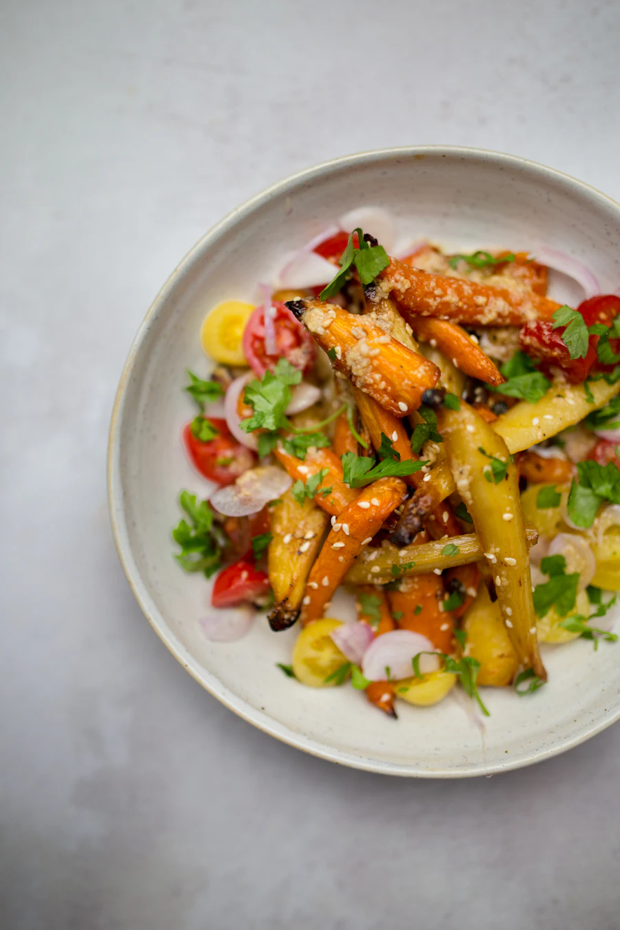 Shallow bowl of roasted carrots, cherry tomatoes, onion slices, herbs, and sesame seeds on a light surface. The dish is photographed from above with soft natural light.