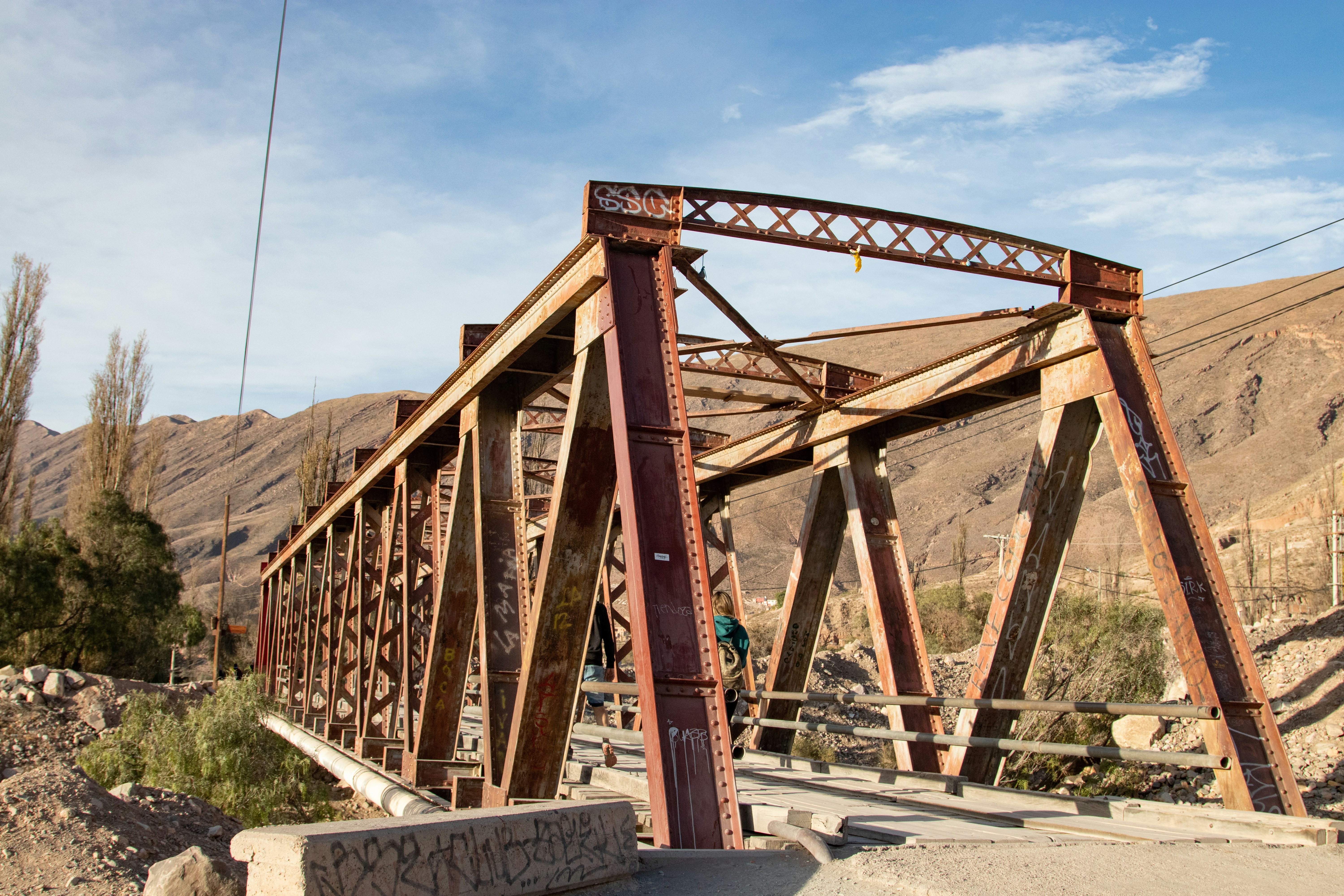 Iron bridge spanning a river against a backdrop of arid mountains and clear blue sky.