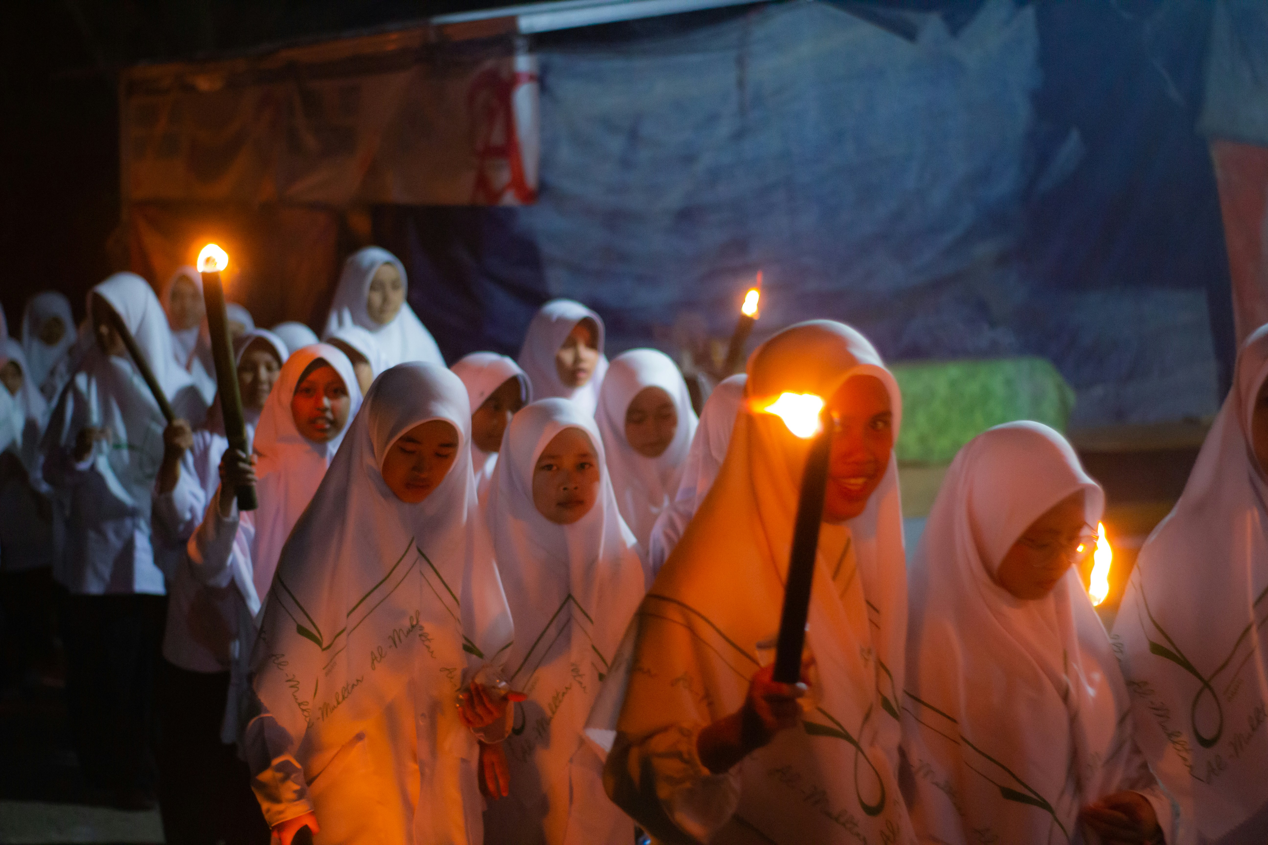 Women in white hijabs hold torches during a night parade, casting warm light on their faces against a dark backdrop.