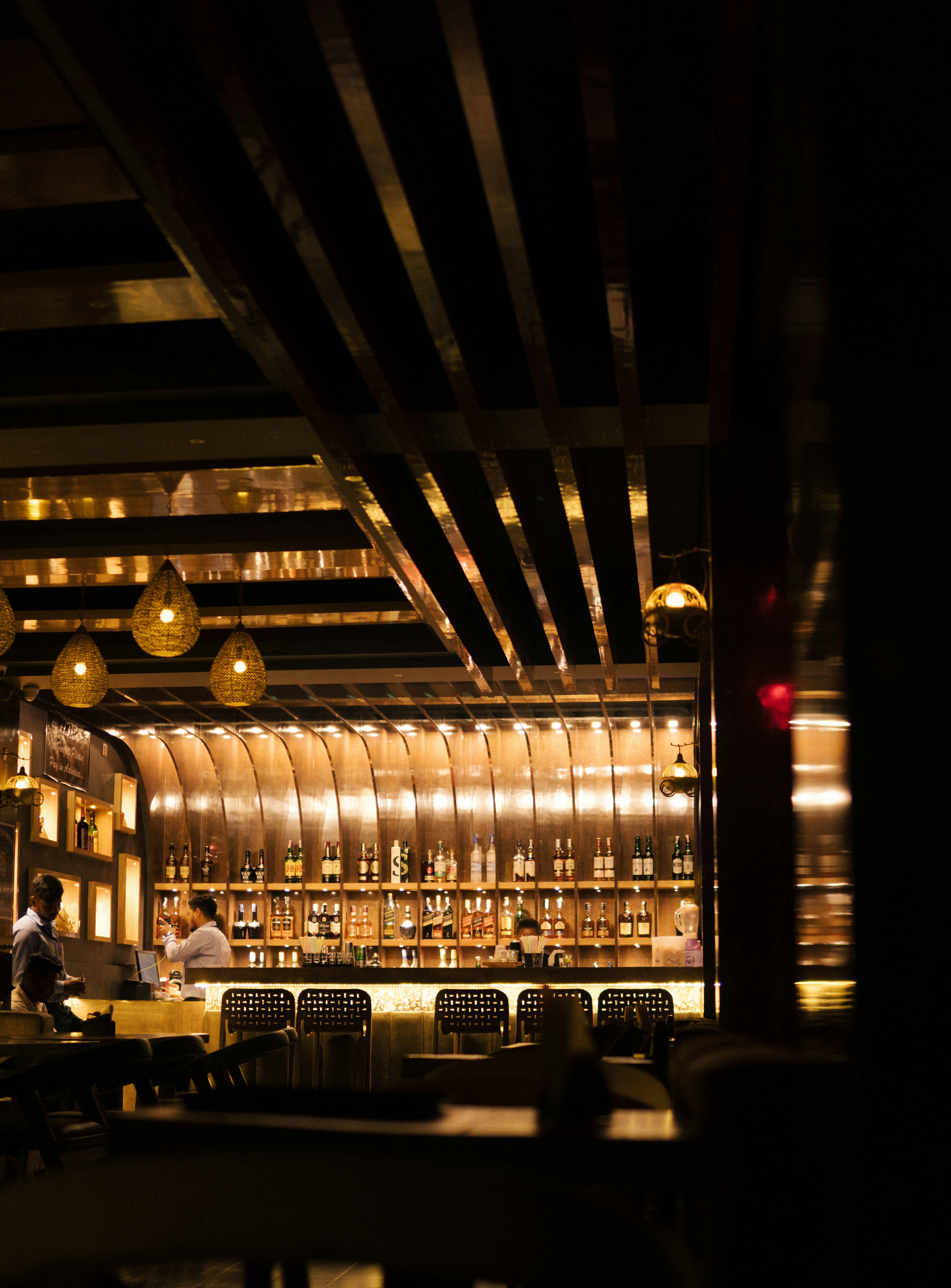 A chic bar interior featuring warm lighting, wooden beams, and a well-stocked shelf of spirits. The ambiance invites patrons to unwind and enjoy.