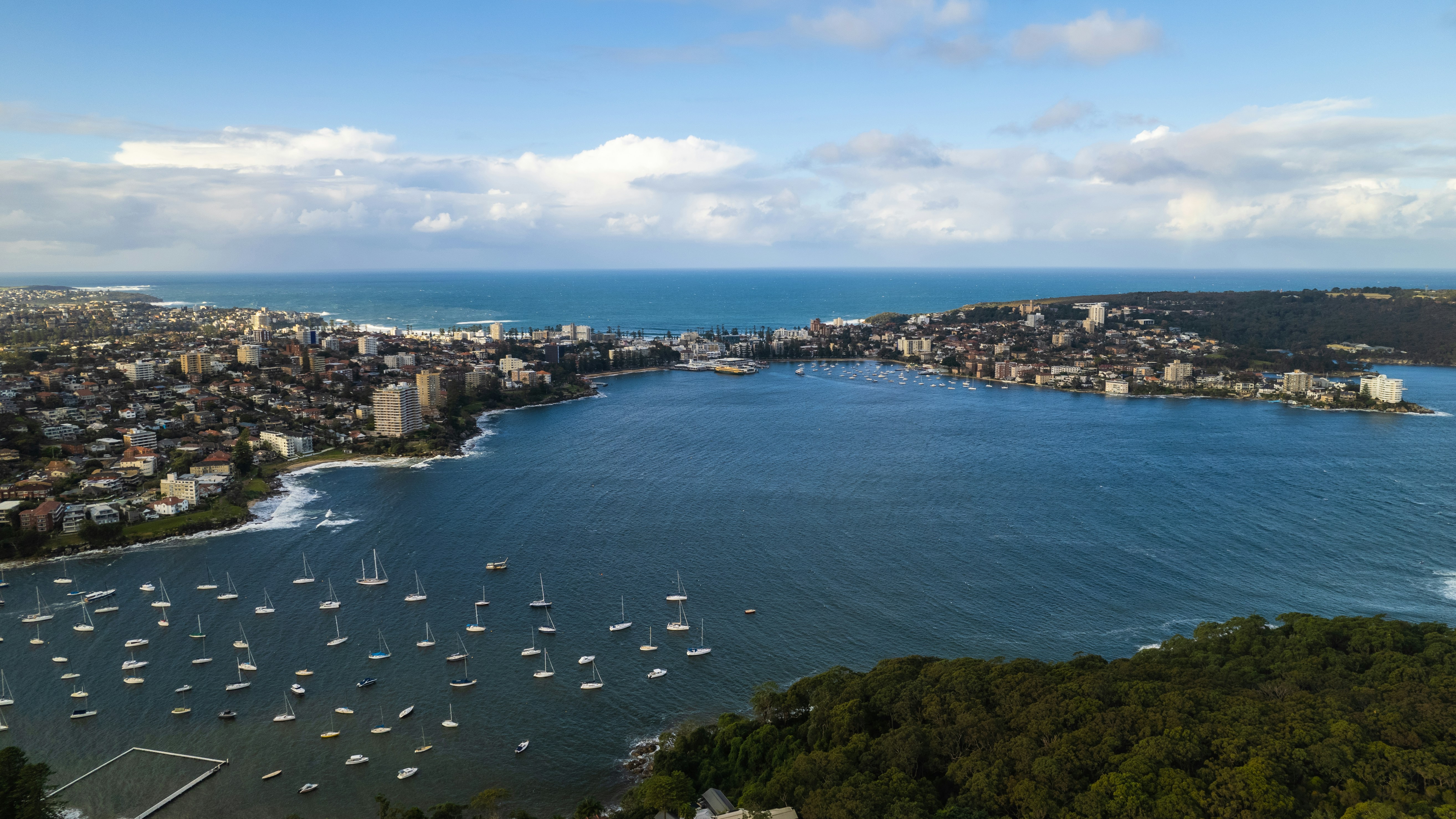 Aerial view of a bustling harbor dotted with sailboats, framed by lush greenery and urban landscapes under a bright sky.