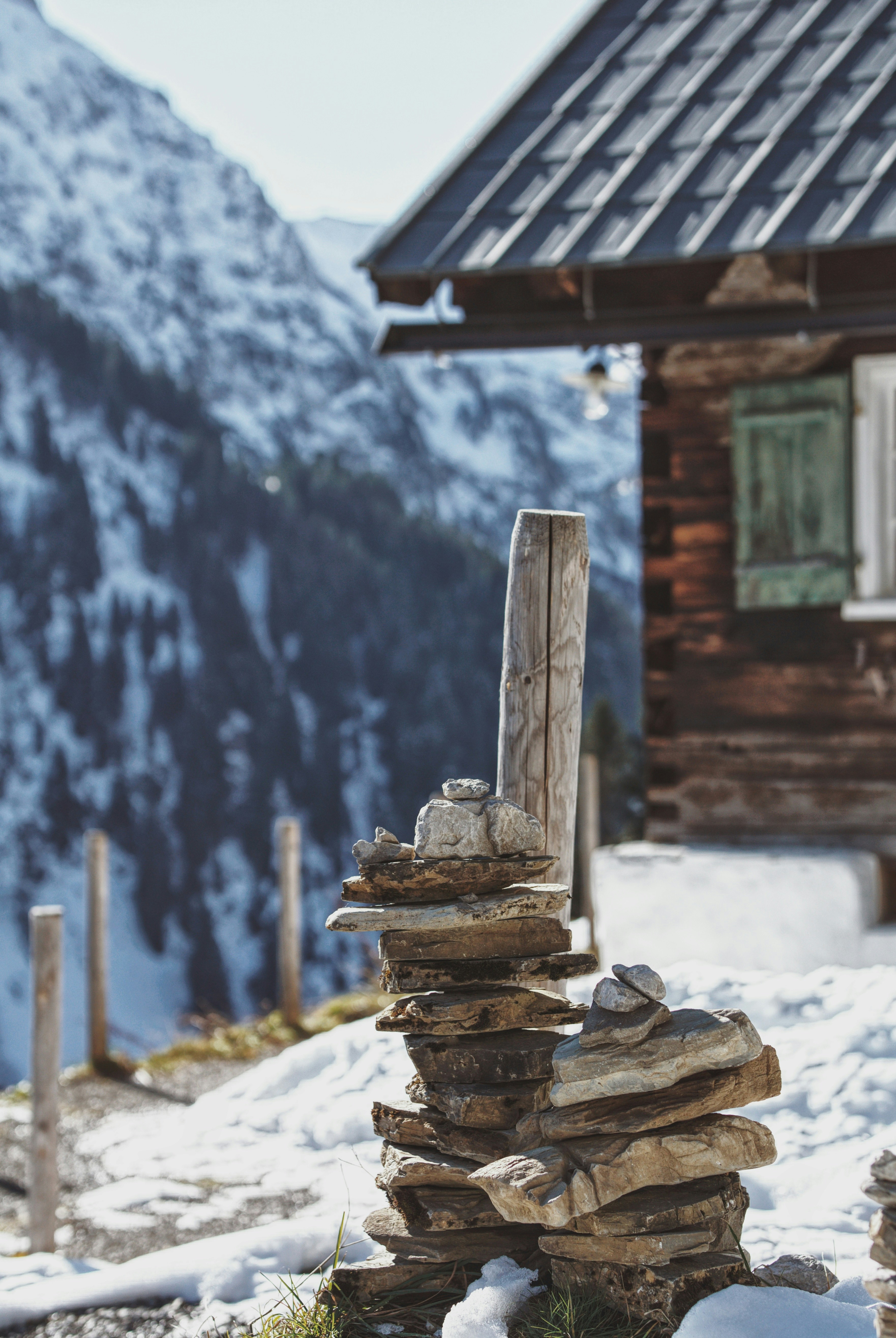 A stack of carefully balanced stones stands in the foreground, with a rustic cabin and snow-covered mountains in the background.