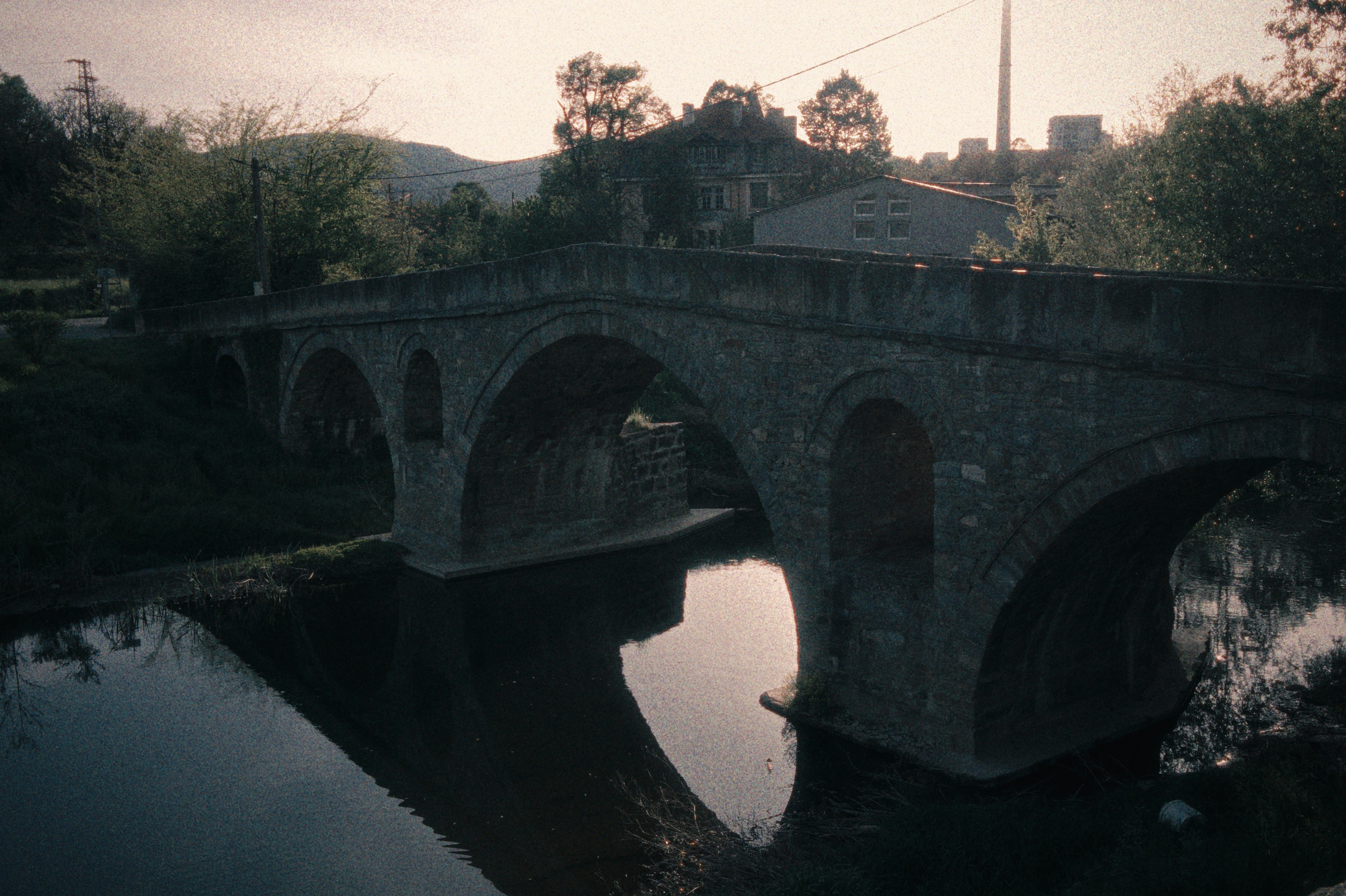 A historic stone bridge arches gracefully over a reflective river, framed by lush greenery and distant buildings. The scene captures a serene moment in nature.