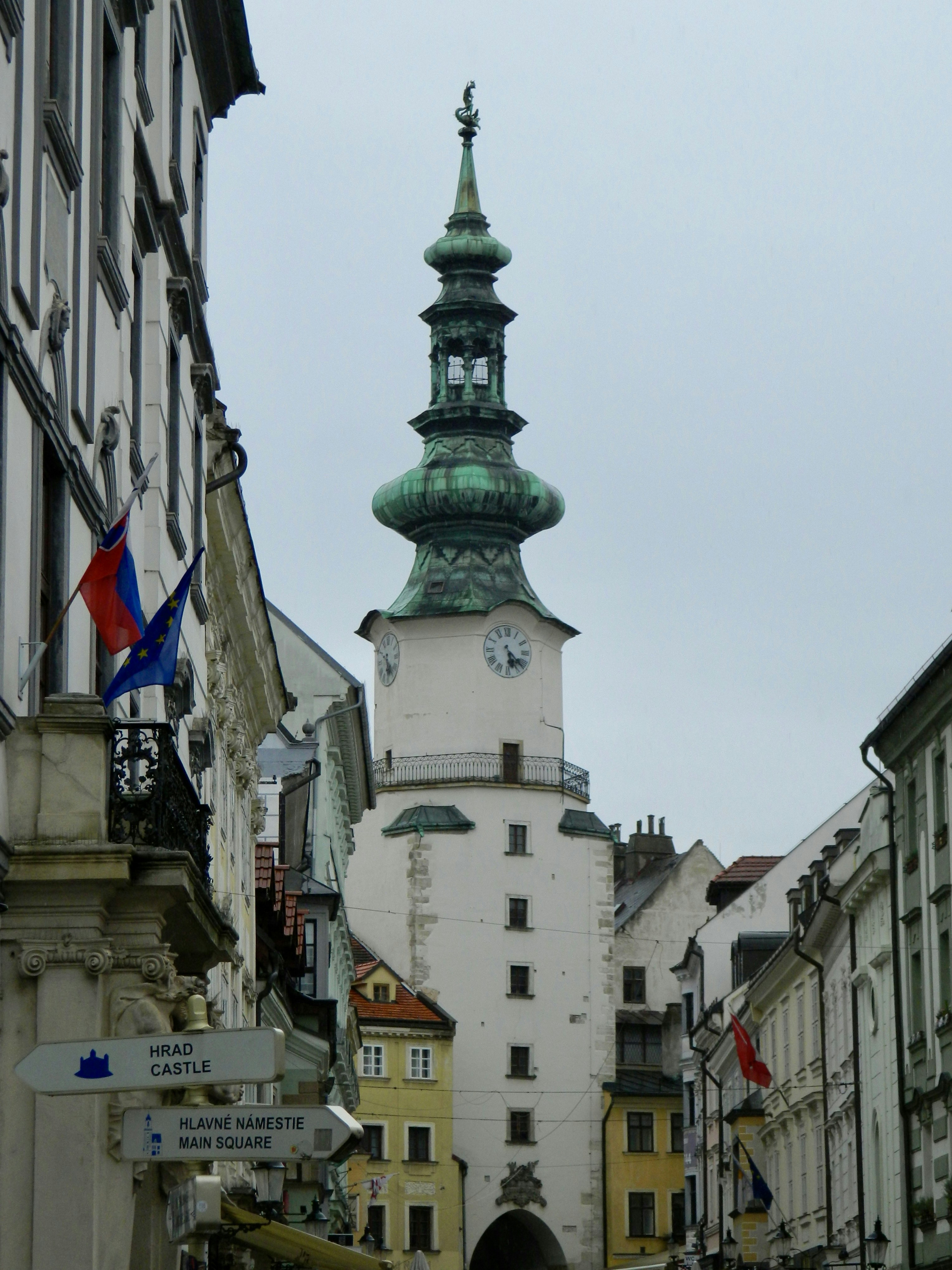 Historic tower adorned with a green dome, surrounded by charming buildings and flags in a quaint street setting.