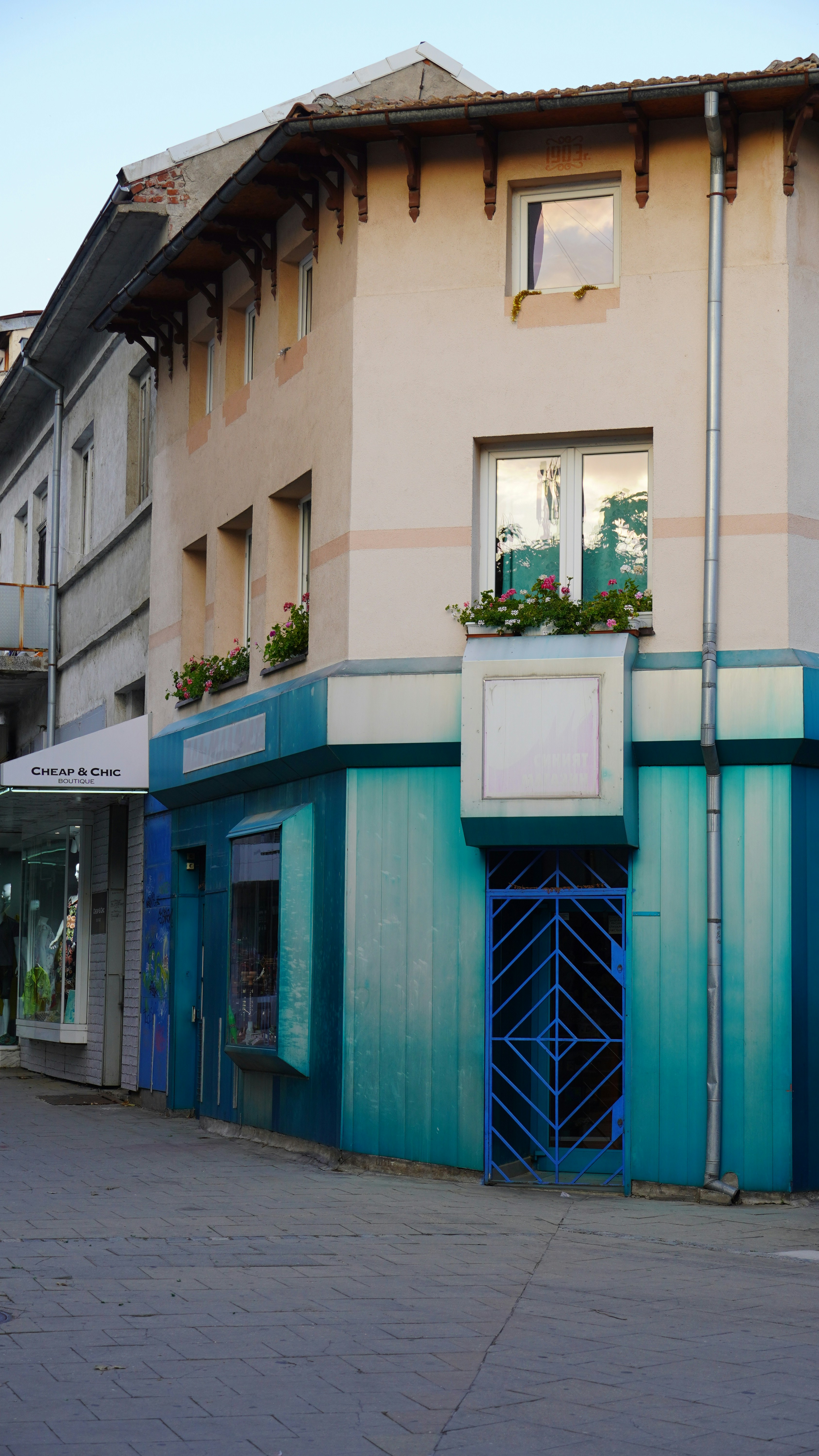 Vibrant blue and green facade of a building adorned with flowers, juxtaposed against a gray structure in an urban setting. The scene captures the essence of city life.