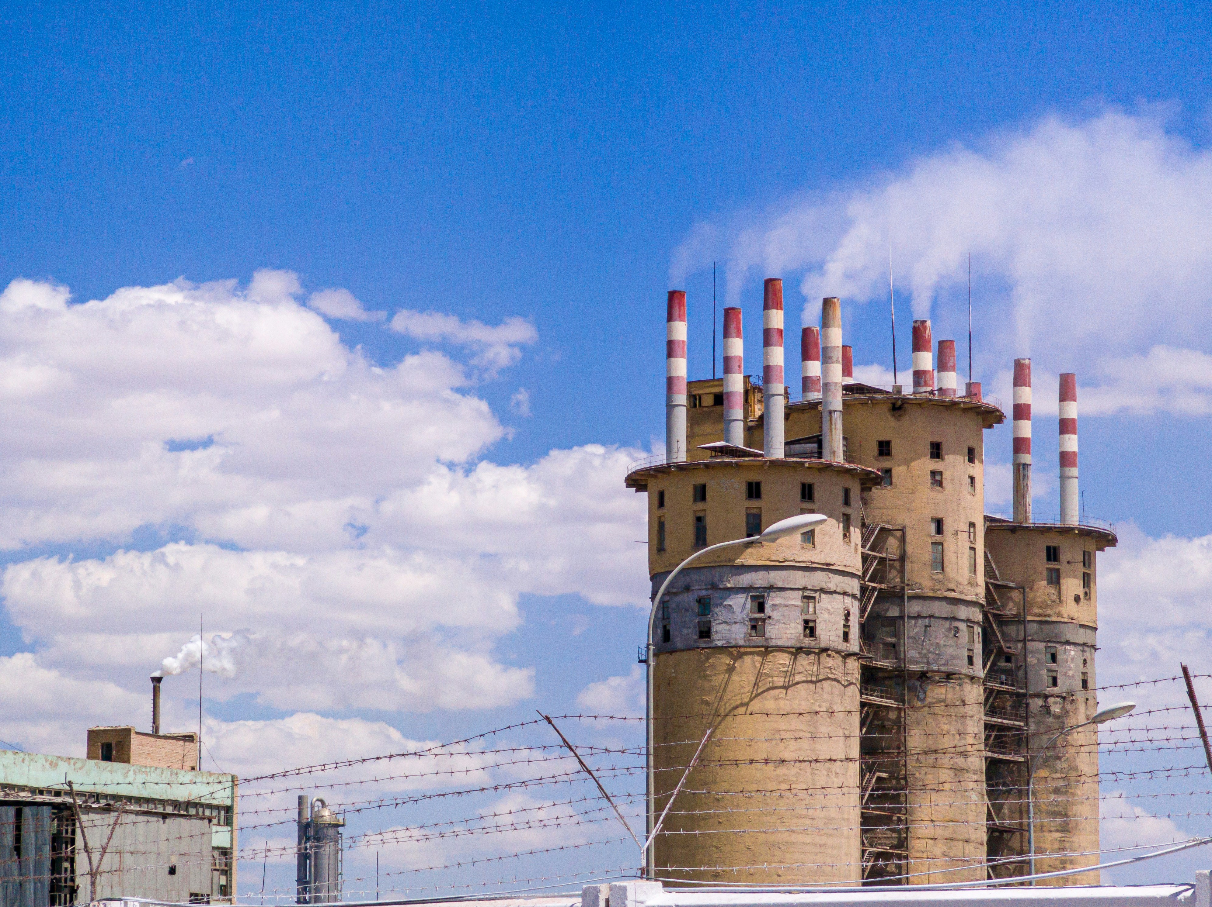 Three towering smokestacks rise above an industrial complex, framed by a bright blue sky dotted with clouds. The scene captures the essence of industrial architecture.