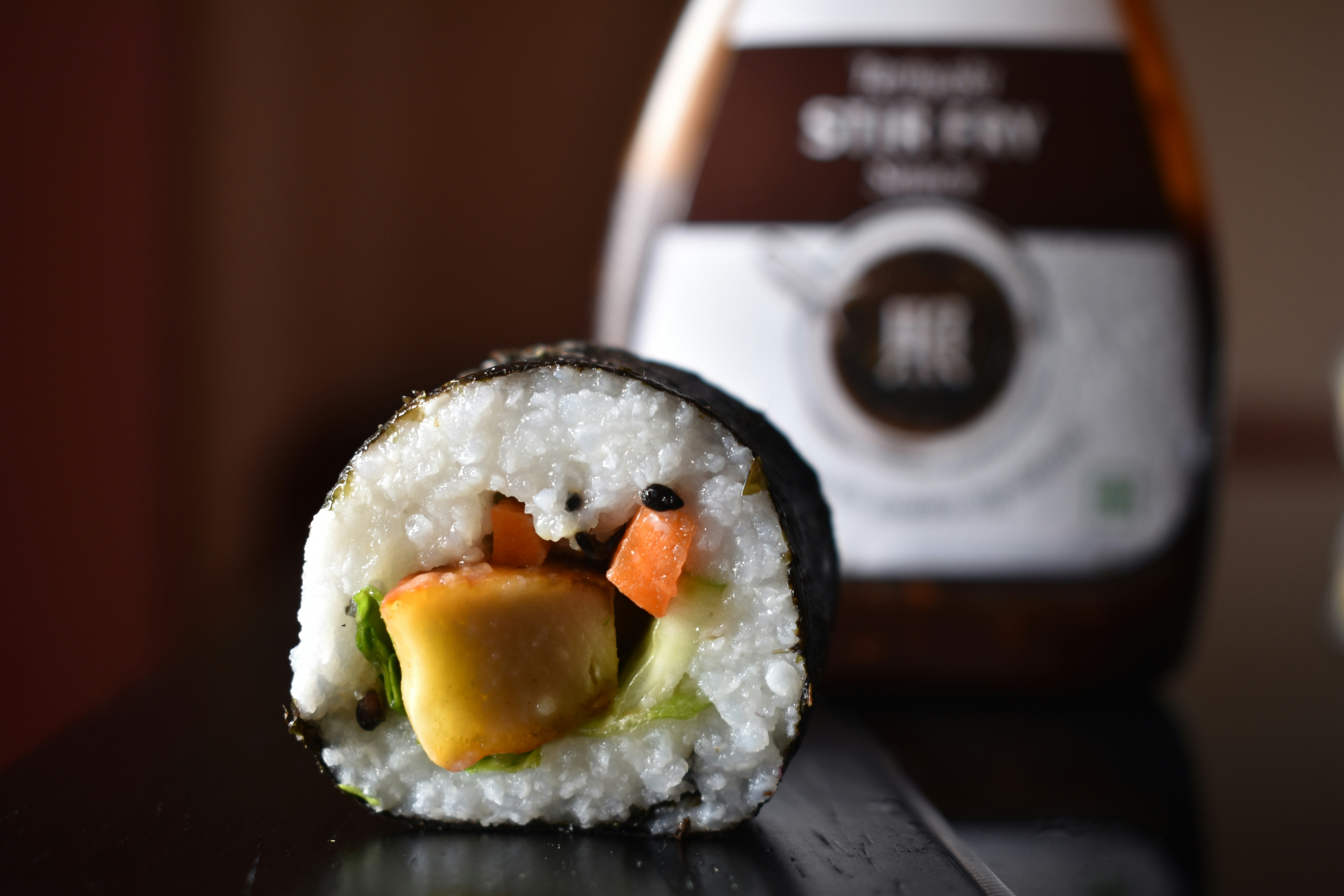 Close-up of a sushi roll showcasing vibrant fillings of vegetables and fruit, with a bottle of sauce blurred in the background.