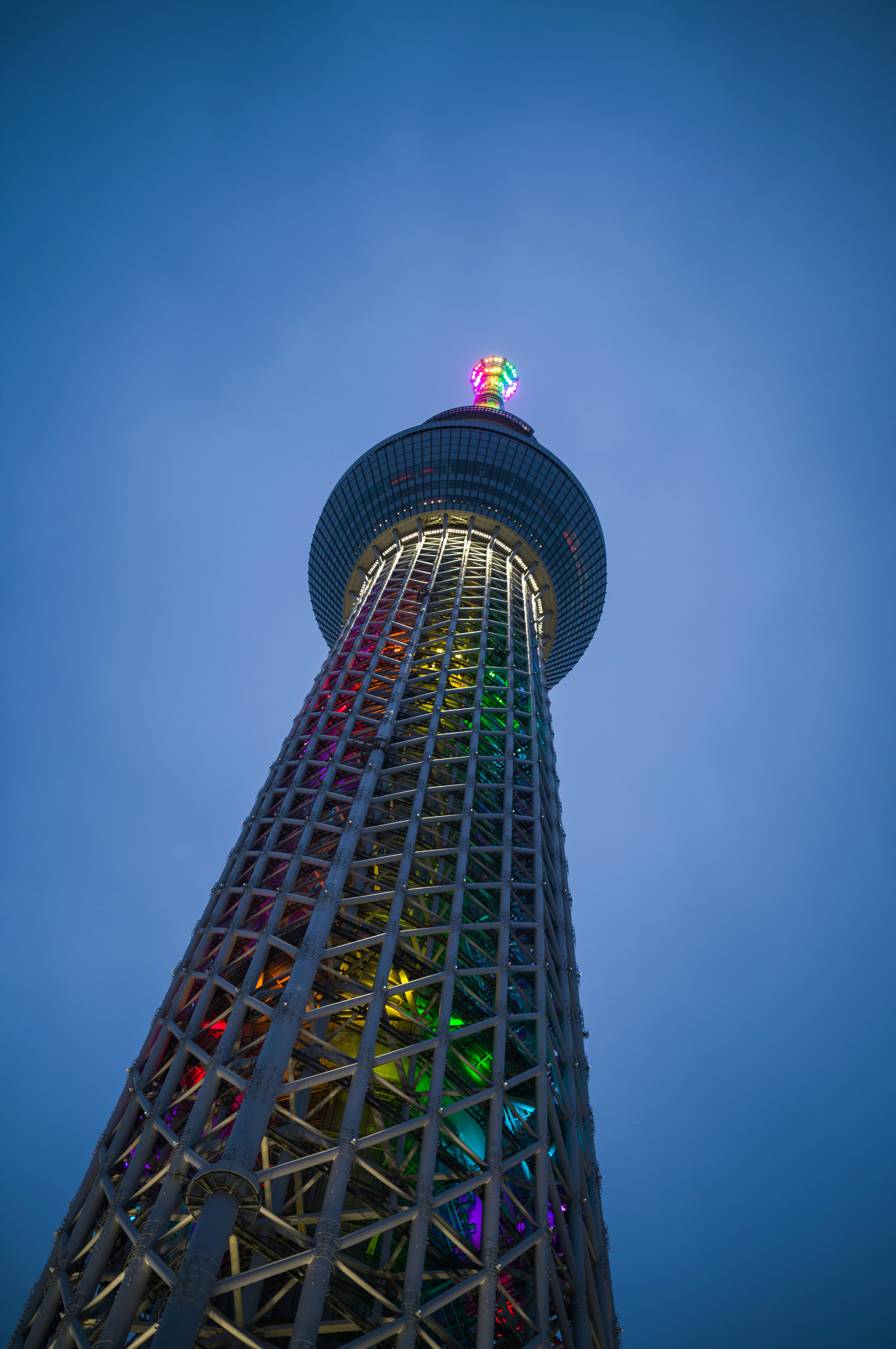 Low-angle photograph of a rainbow-lit lattice tower rising into a dusk-blue sky, featuring a multicolored observation deck and a lit spherical beacon at the summit.
