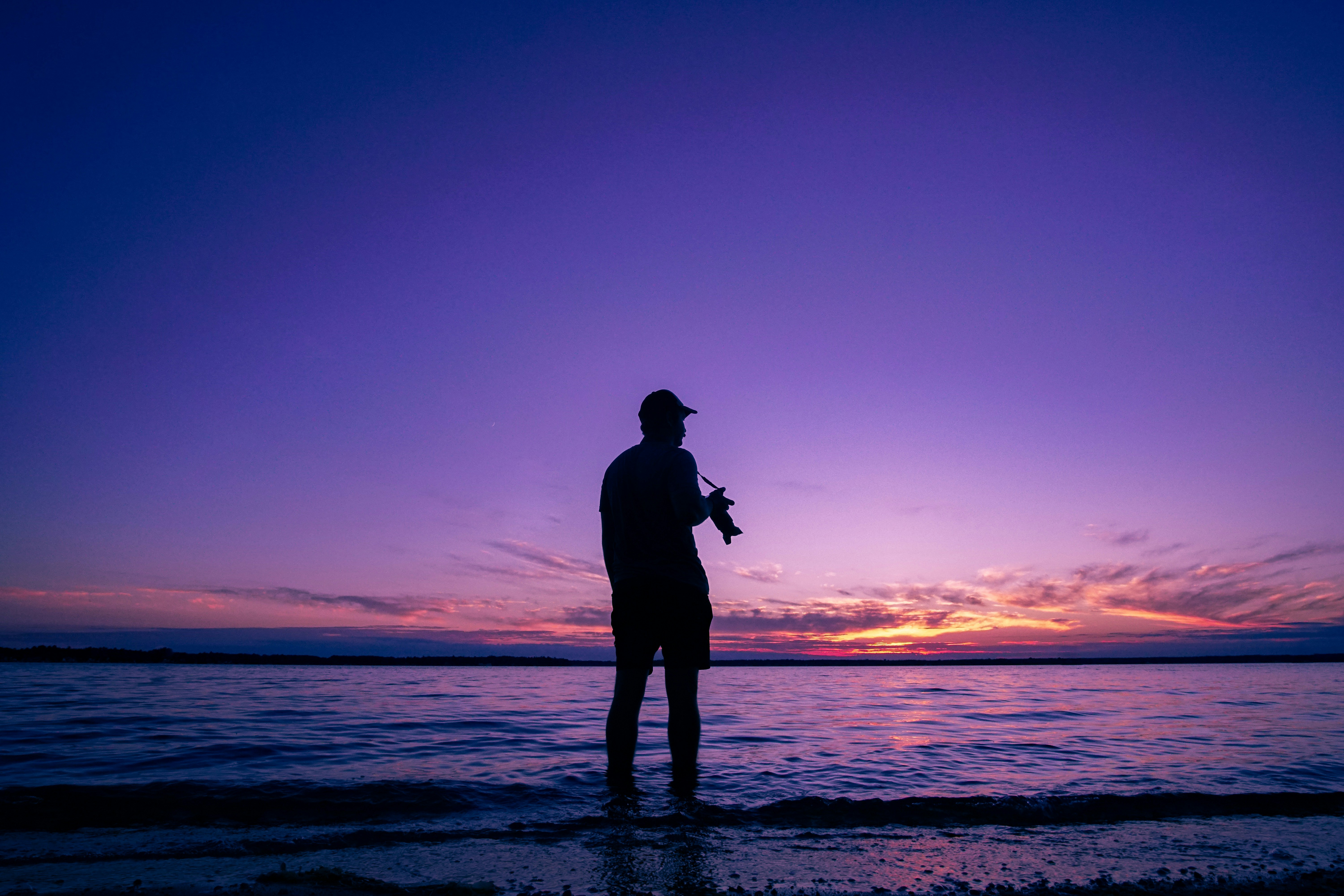 A silhouetted figure stands in shallow water, holding a camera, against a vibrant purple and orange sunset sky. The scene captures the tranquility of twilight moments by the shore.