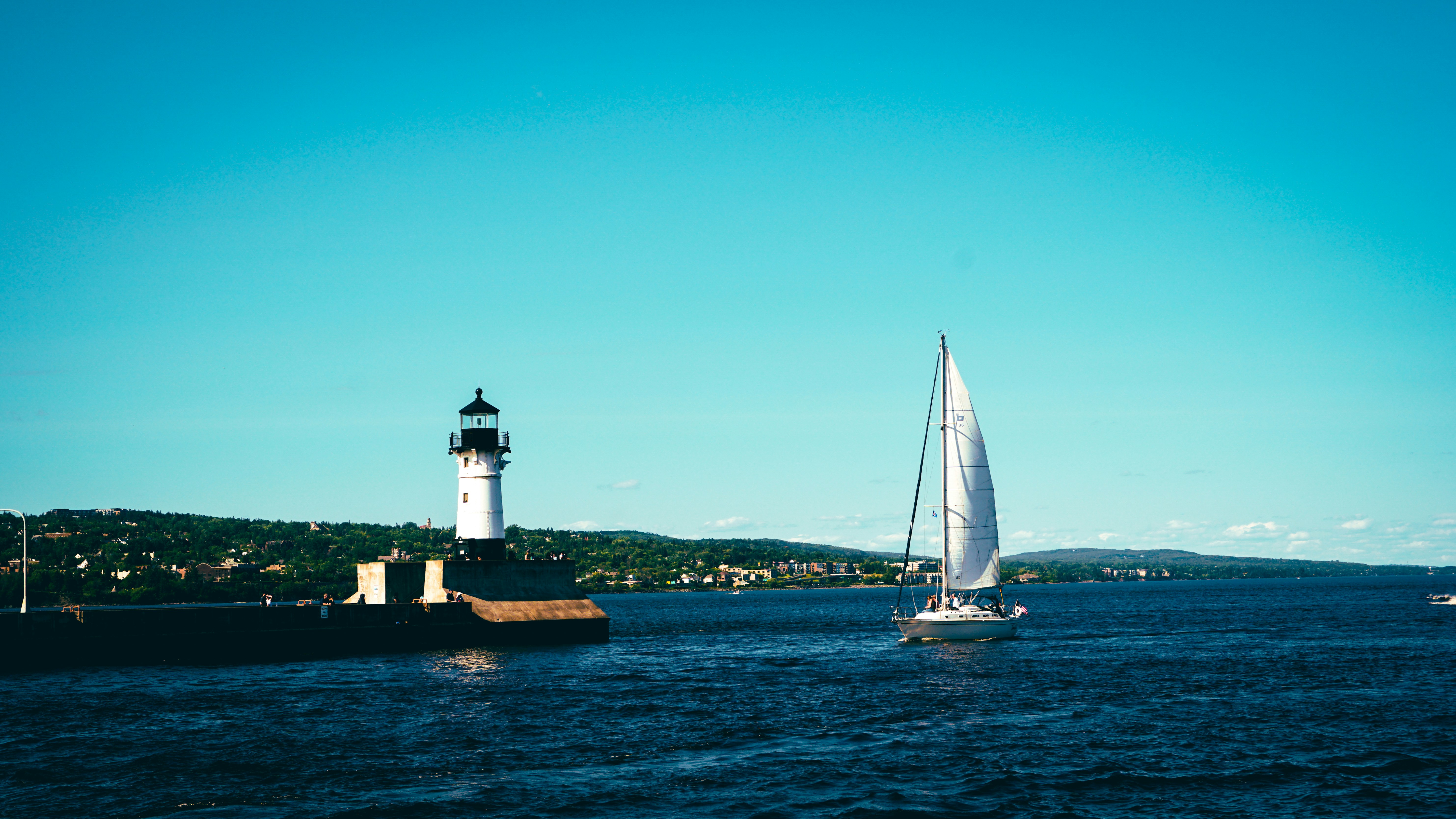Sailboat gliding near a lighthouse on a clear day with distant hills.