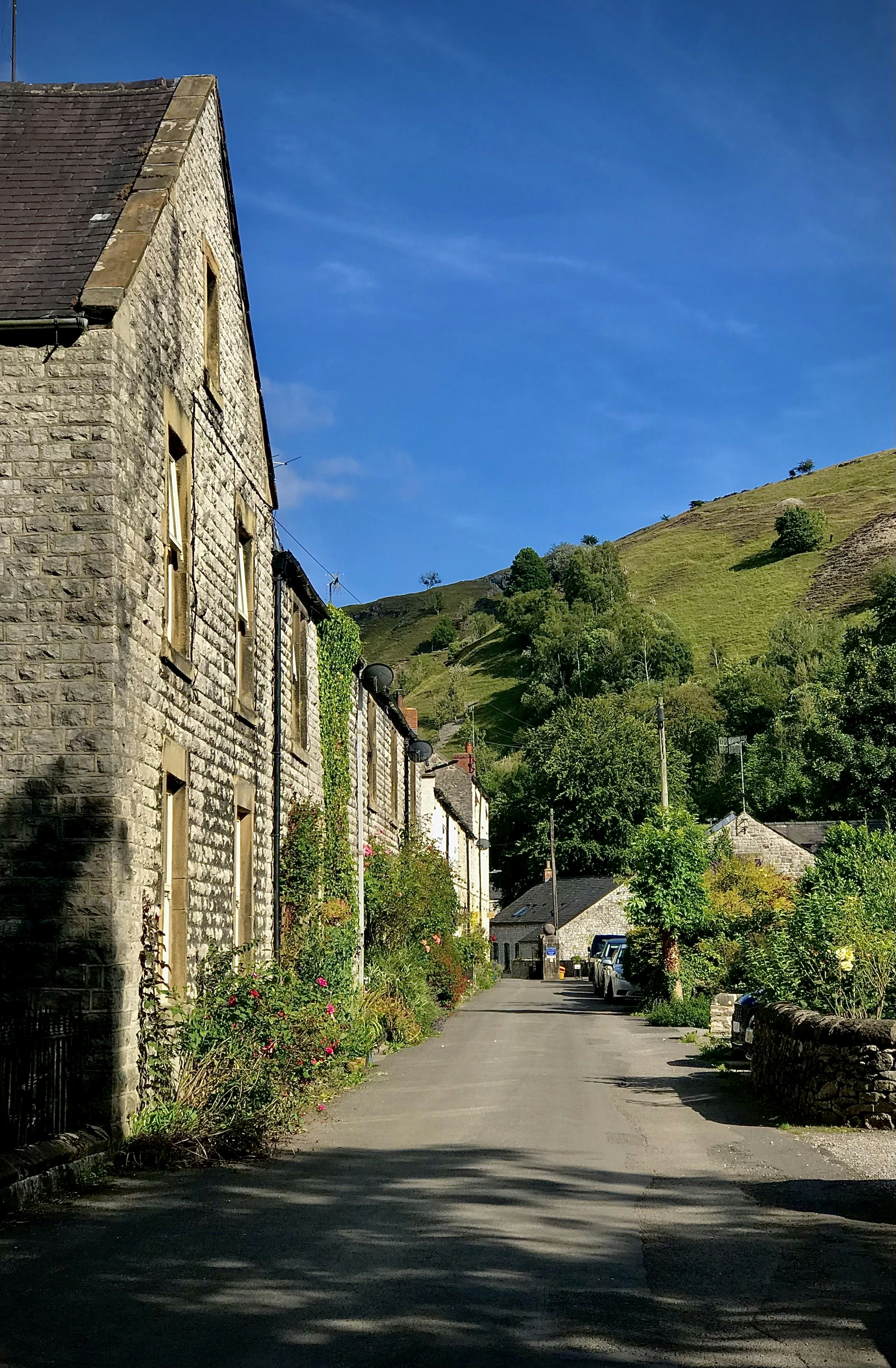 Charming stone houses line a narrow village lane, flanked by lush greenery and rolling hills under a bright blue sky.