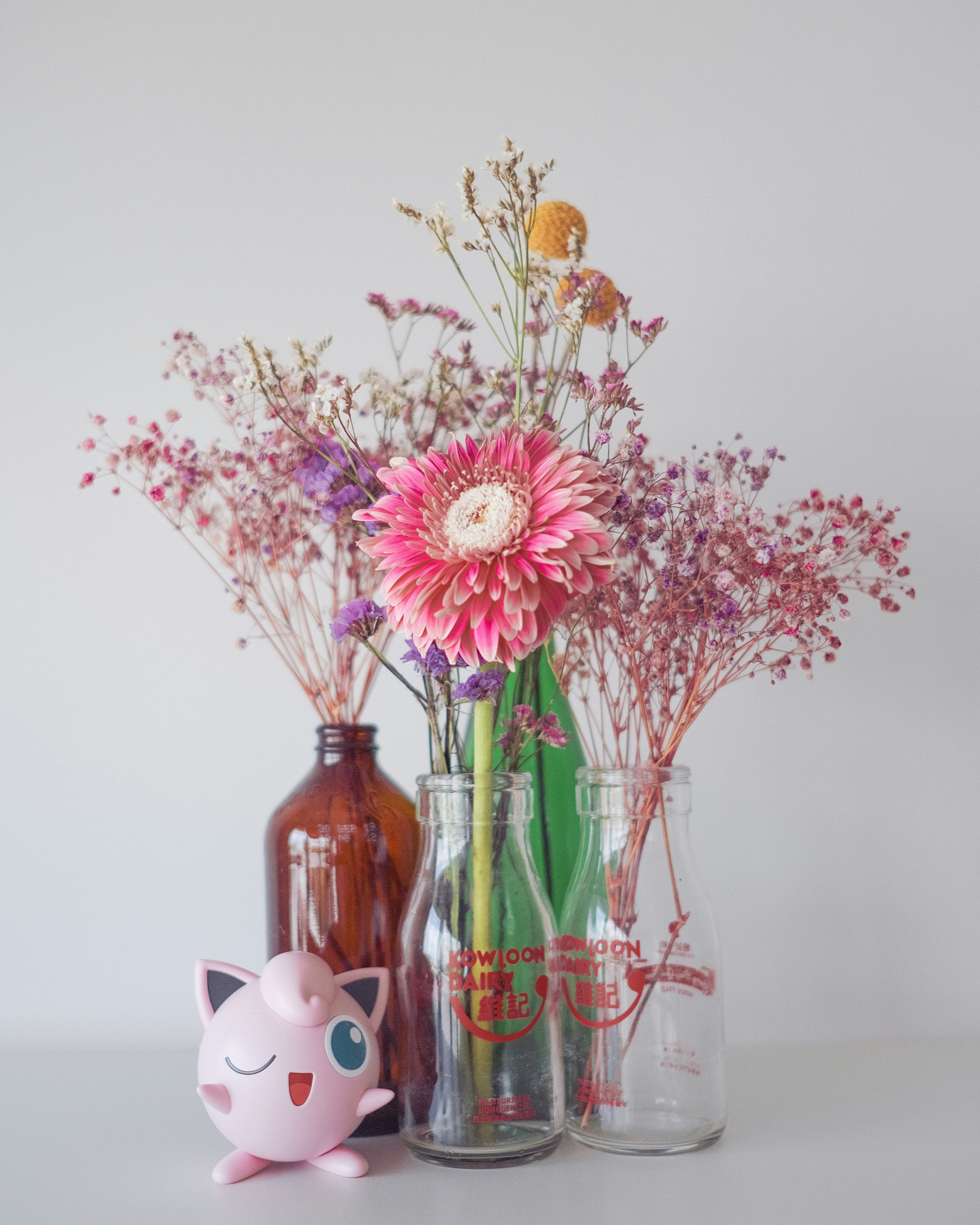 A playful arrangement of vibrant flowers in glass bottles, featuring a pink Jigglypuff figurine in the foreground.