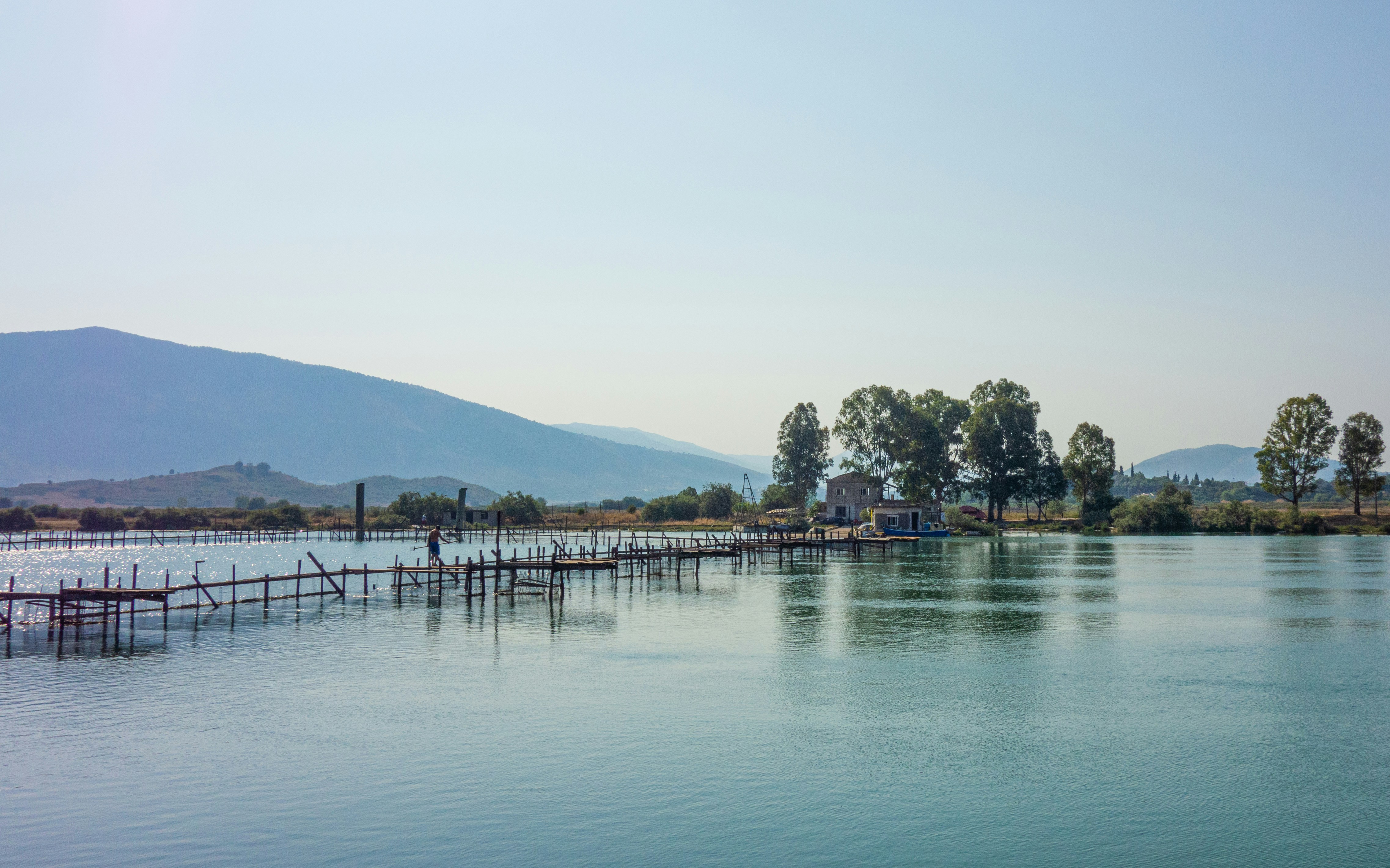 A tranquil lakeside scene featuring a wooden pier extending into calm waters, surrounded by lush trees and distant mountains. The clear sky enhances the serene atmosphere.