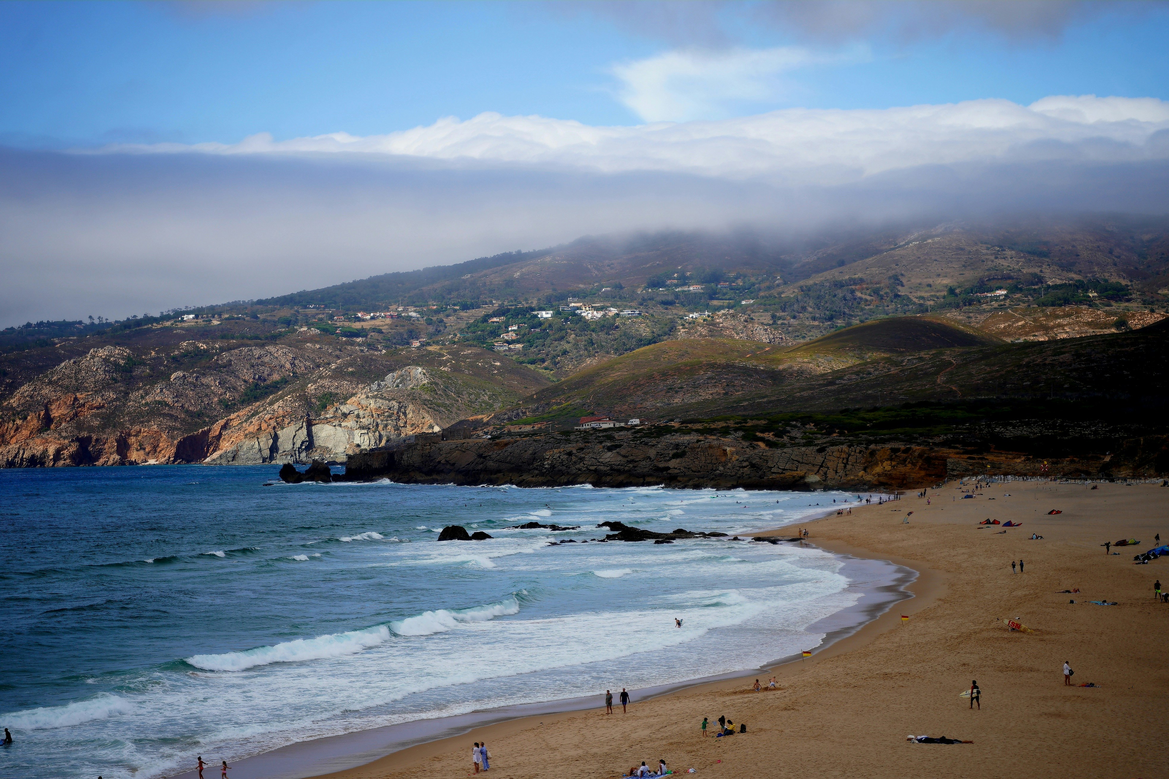 Expansive beach scene with gentle waves lapping at the shore, framed by rolling hills and a cloudy sky.