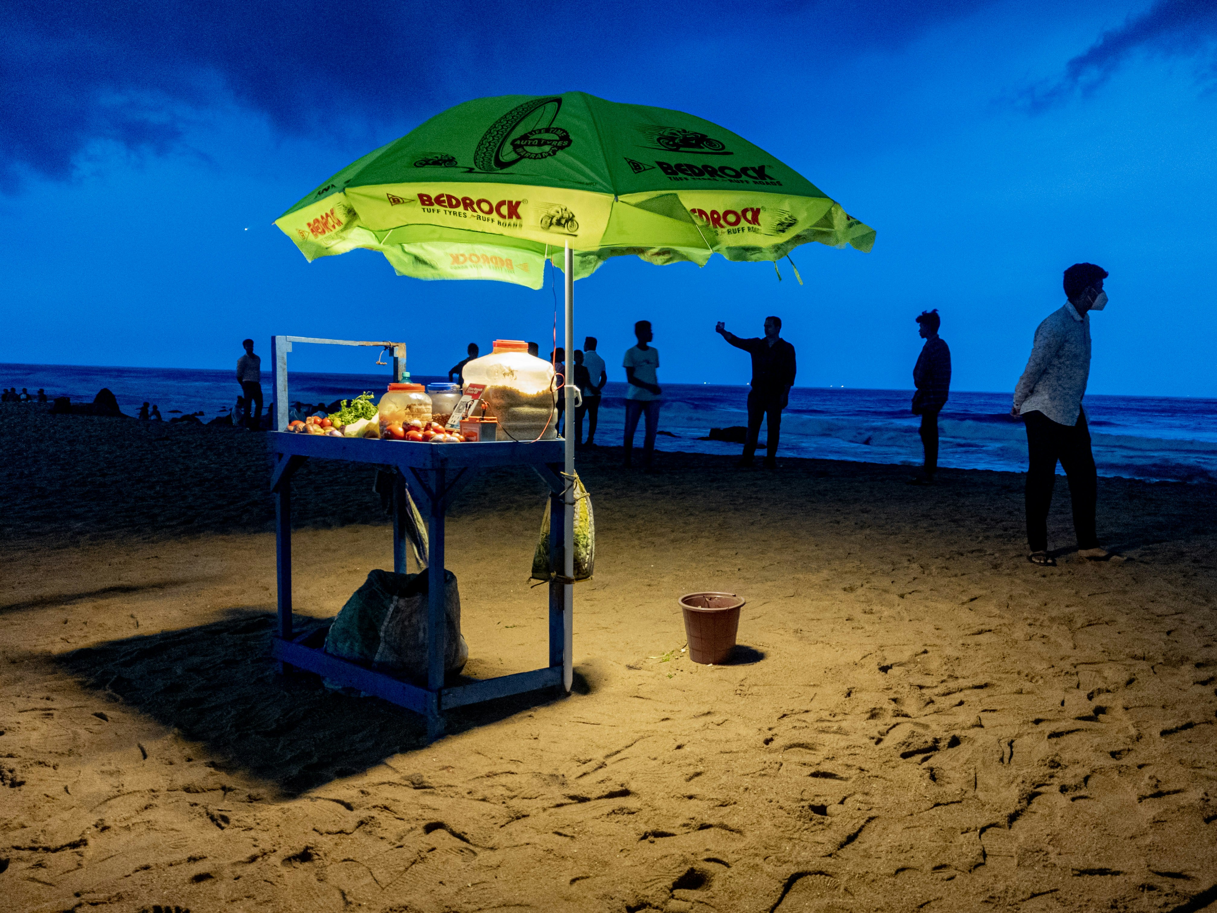Beachside food cart illuminated by a green umbrella, with silhouettes of people against a twilight ocean backdrop.