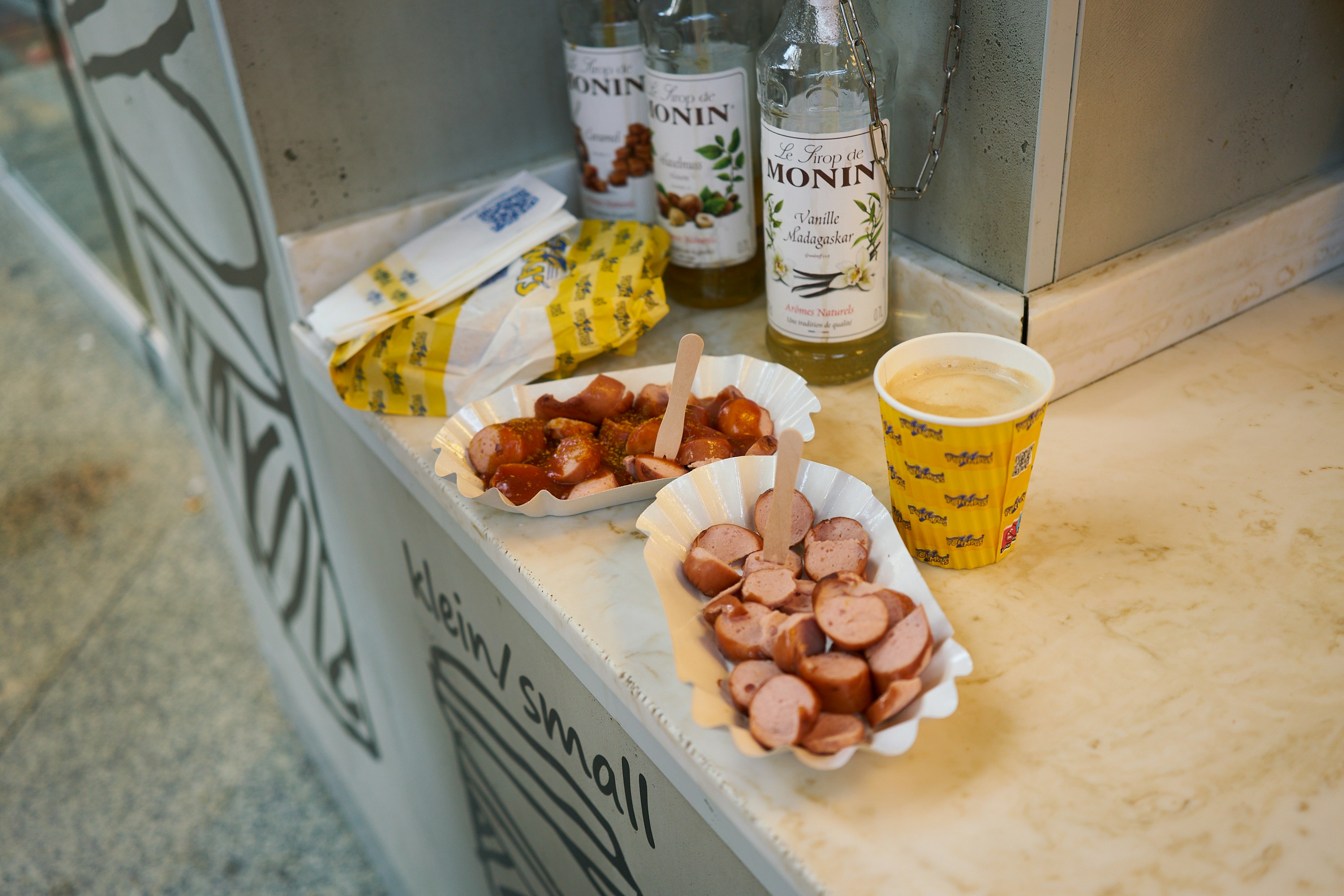 Plates of sliced sausages and skewered bites alongside a cup of beverage, set against a textured background. The scene captures the essence of casual street dining.