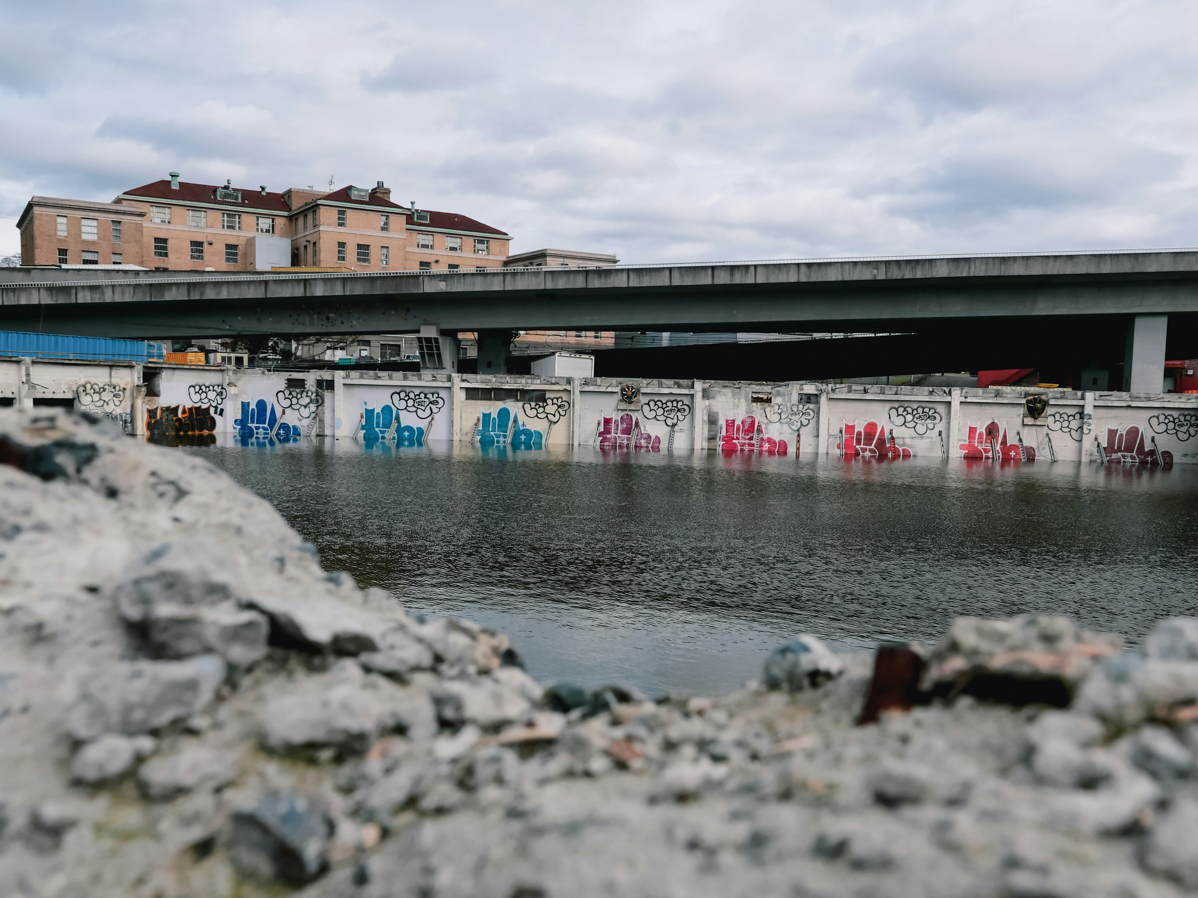 Wide-angle street-scene photograph of a graffiti-covered wall along a calm river, with a concrete bridge overhead and a rocky foreground.