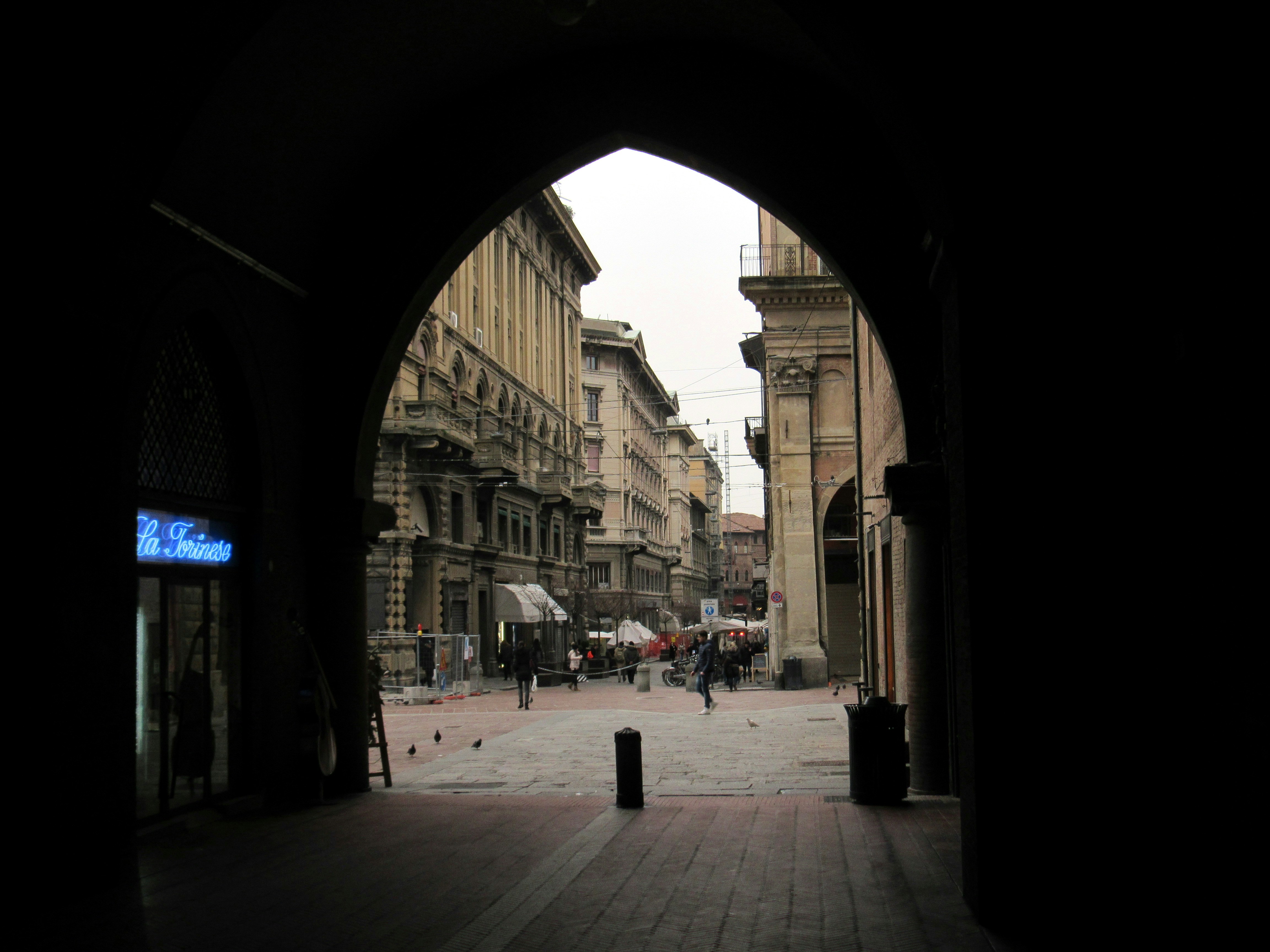 Photograph of a sunlit pedestrian street framed by a dark archway, with umbrellas and pedestrians along historic façades.