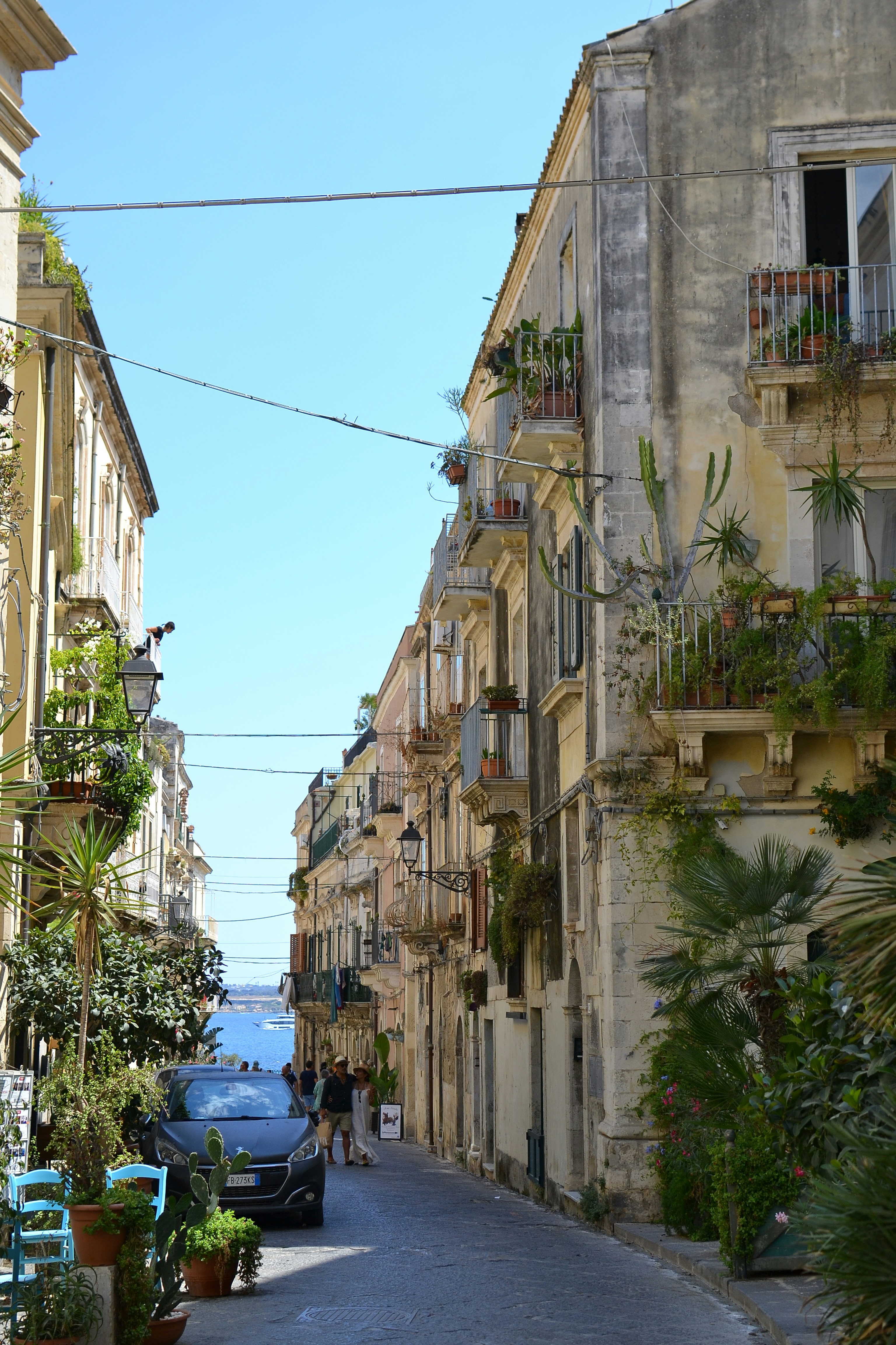 Narrow street lined with vibrant plants and charming buildings, leading to a glimpse of the sea. The scene captures the essence of Mediterranean life.