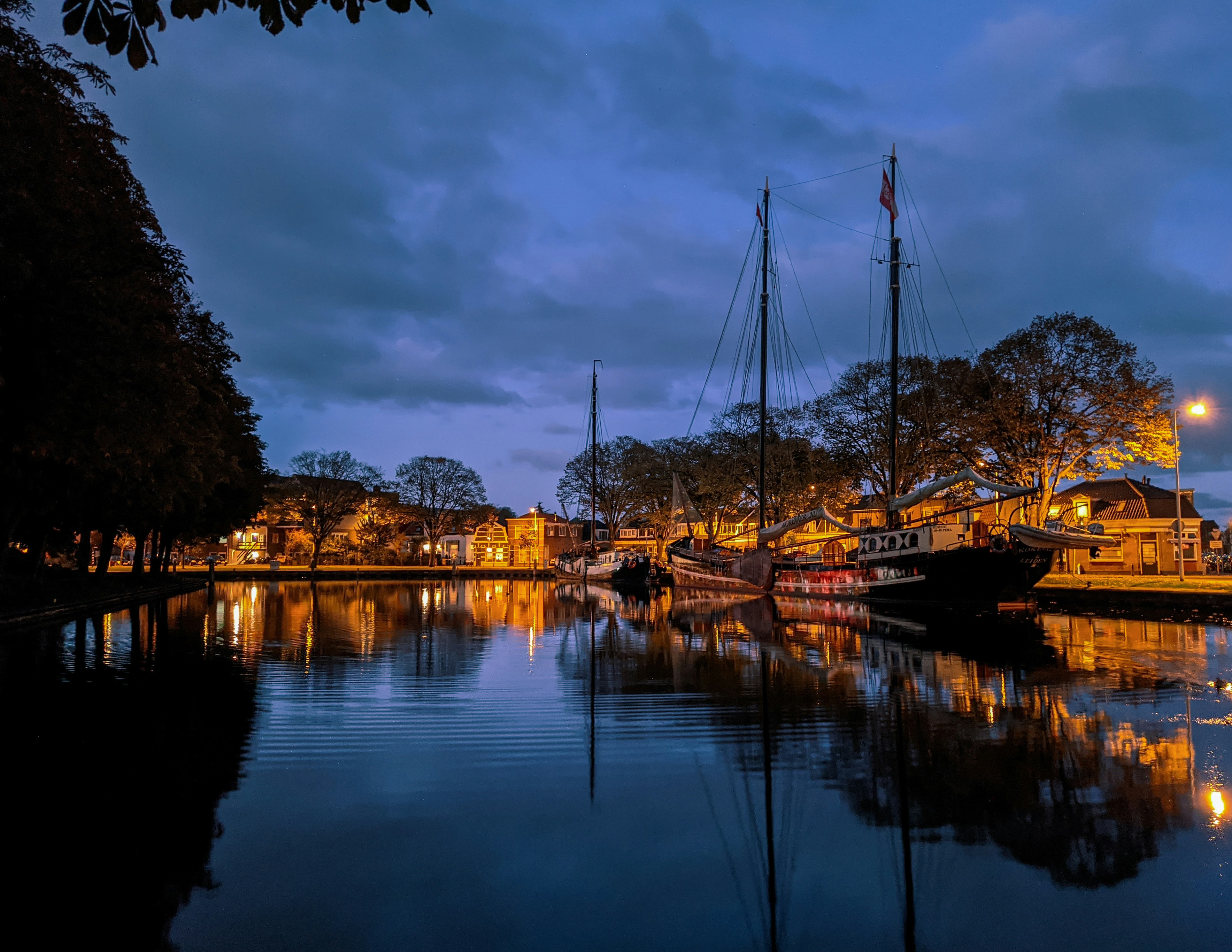 Nighttime harbor scene with sailboats and buildings illuminated by warm lights reflecting on calm water.