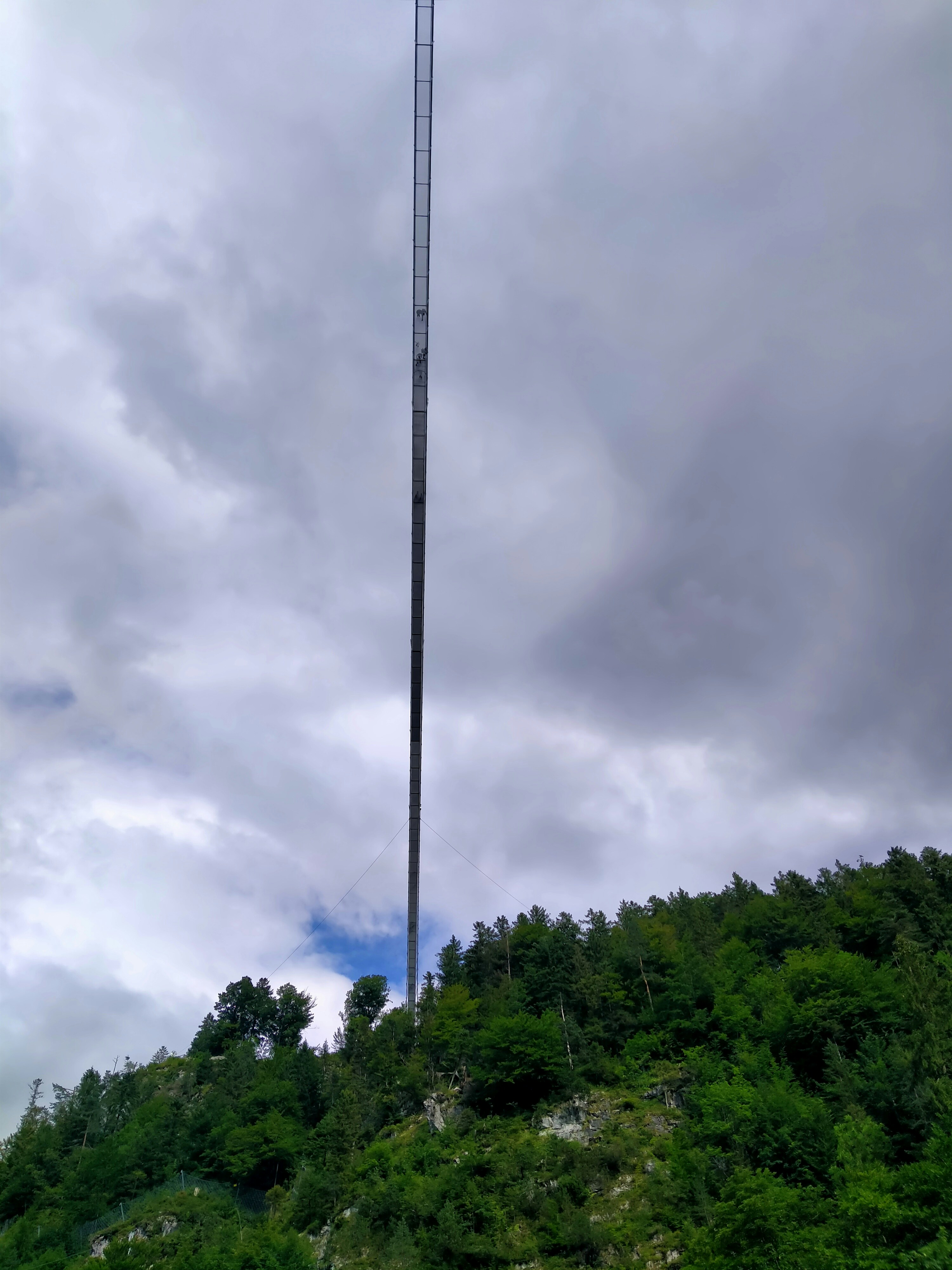 A tall communication tower rises dramatically against a backdrop of dense greenery and overcast skies.