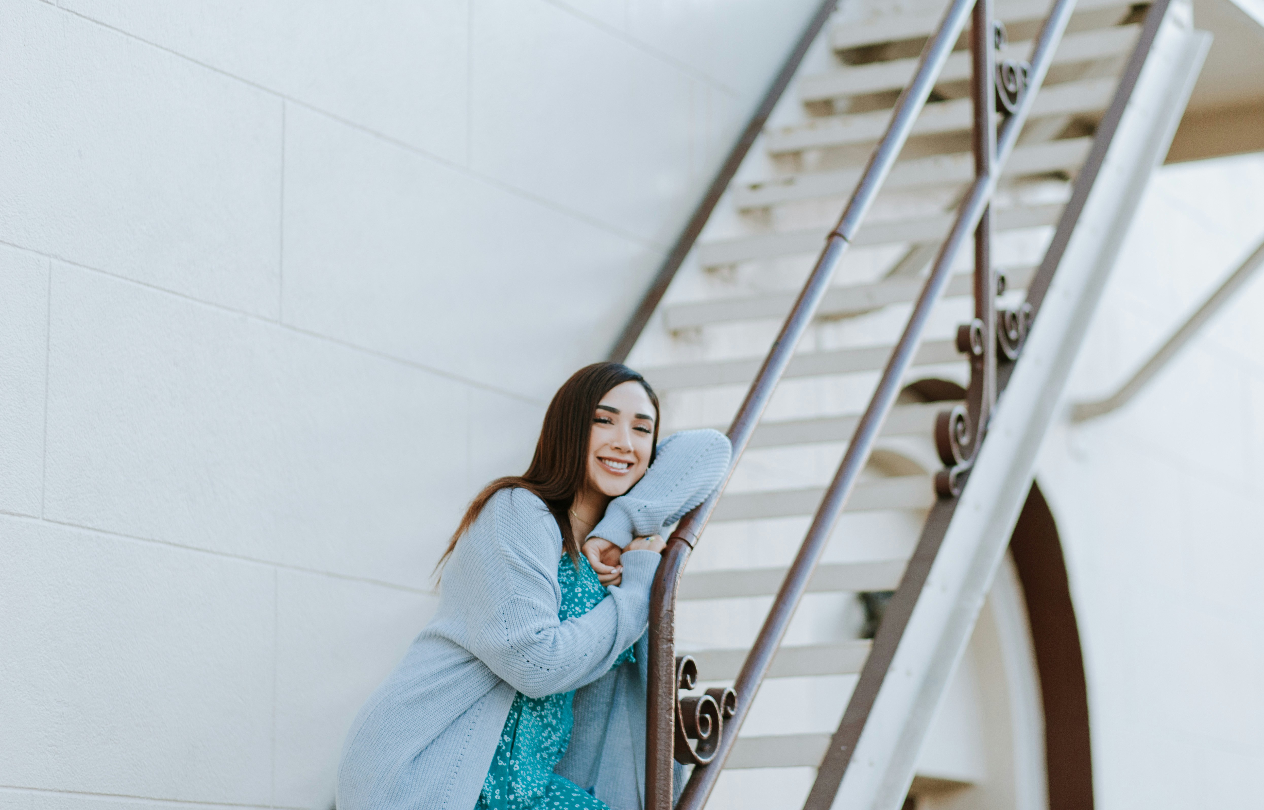 Woman in a blue dress and cardigan leaning against a staircase railing with a peaceful expression.