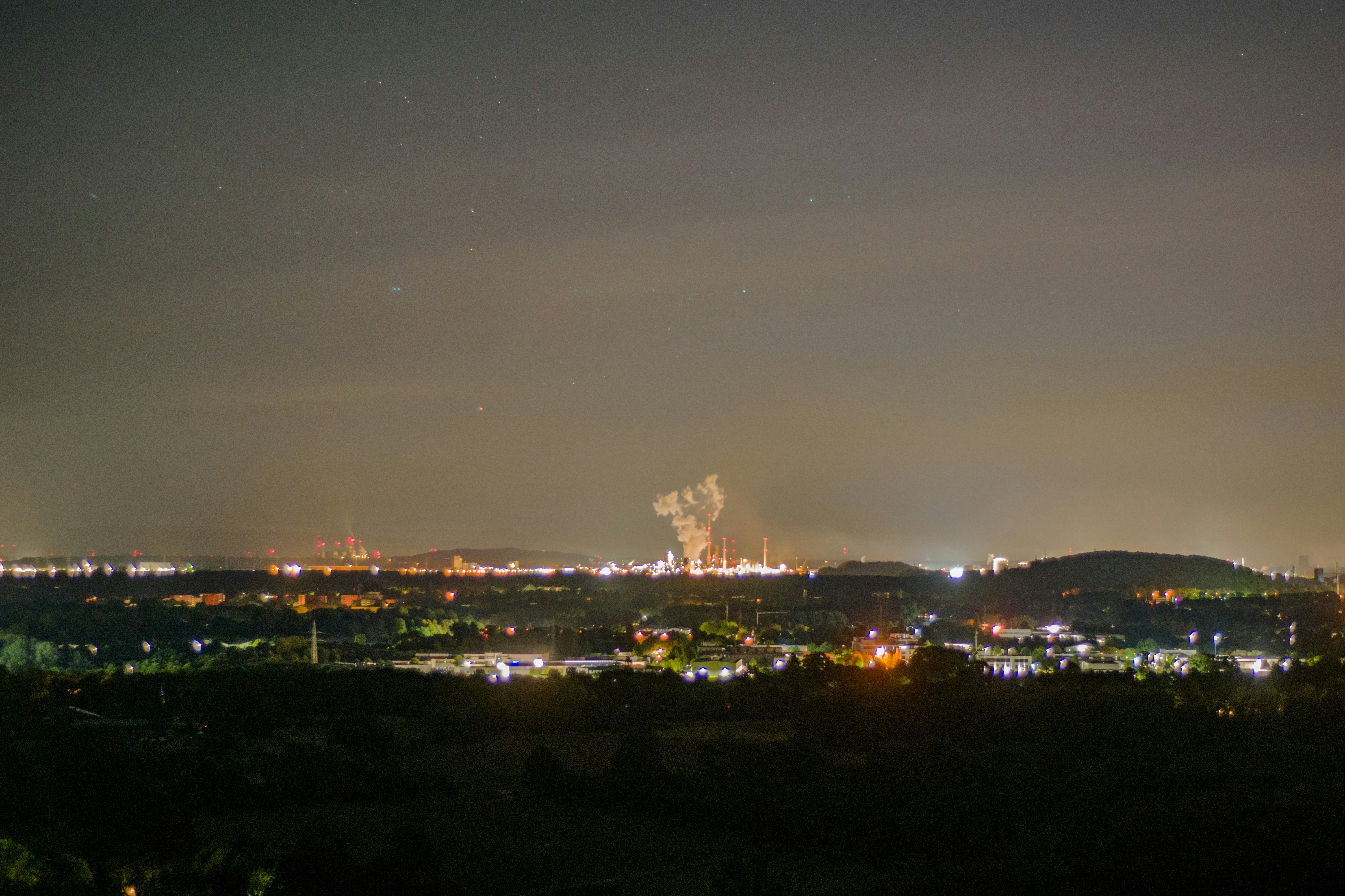 Illuminated skyline of an industrial area at night, featuring distant fireworks amidst a backdrop of stars and clouds.