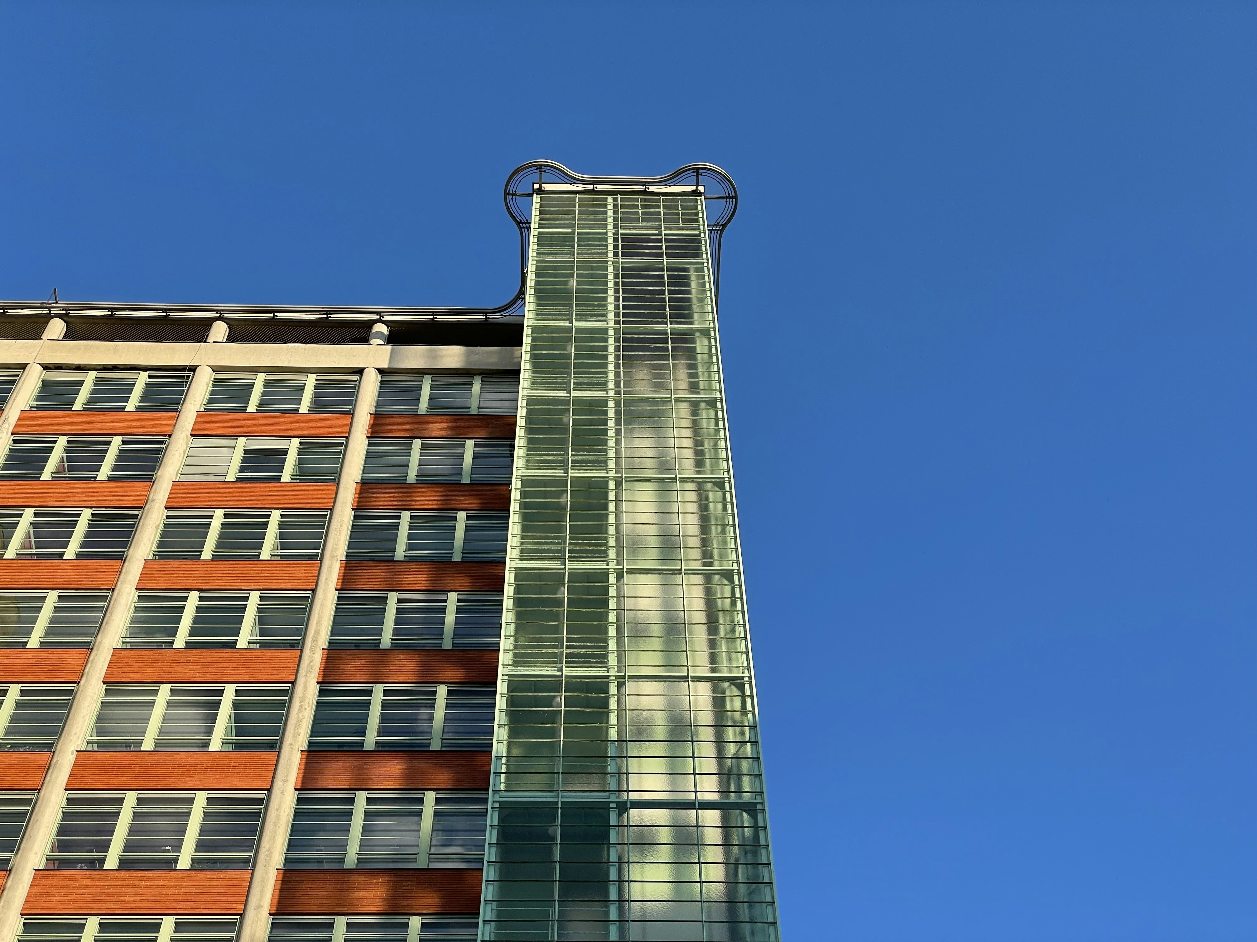 Modern building with orange and white grid facade and glass elevator tower under clear blue sky.