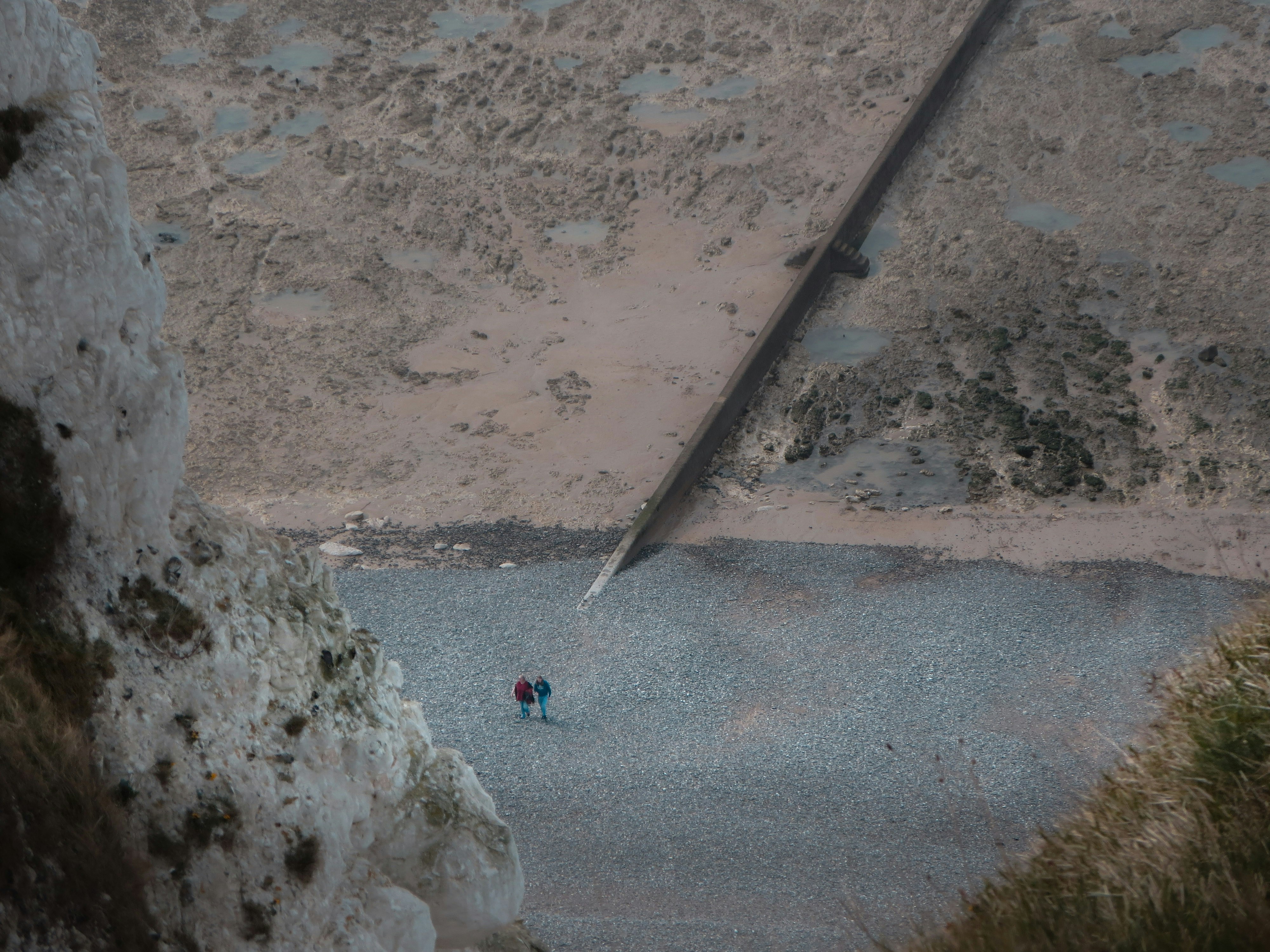 Two figures walking along a pebbled beach beneath towering cliffs, with a striking diagonal line of a structure leading the eye across the scene.