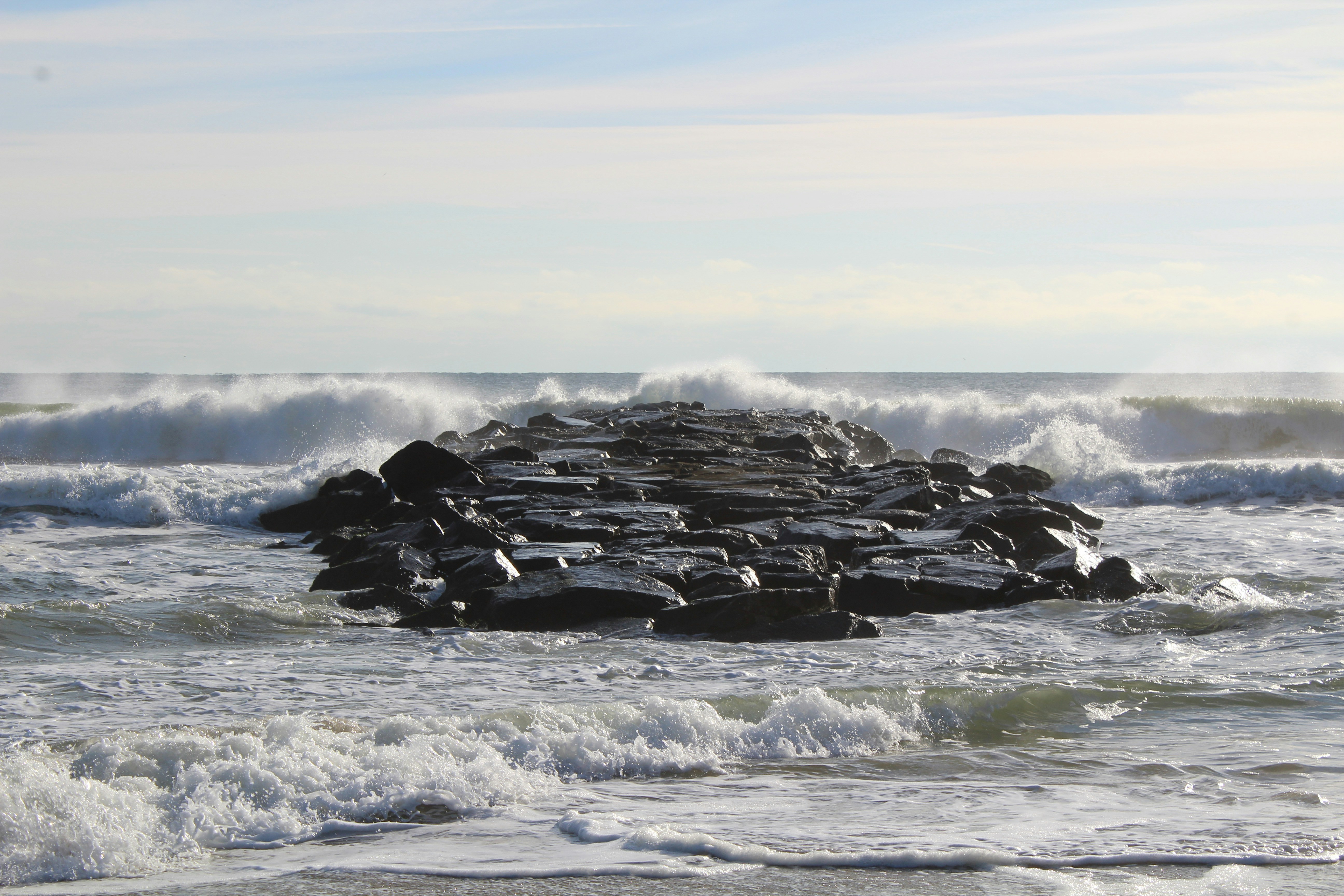 Rocky jetty extending into the ocean with waves crashing over its surface under a bright sky.