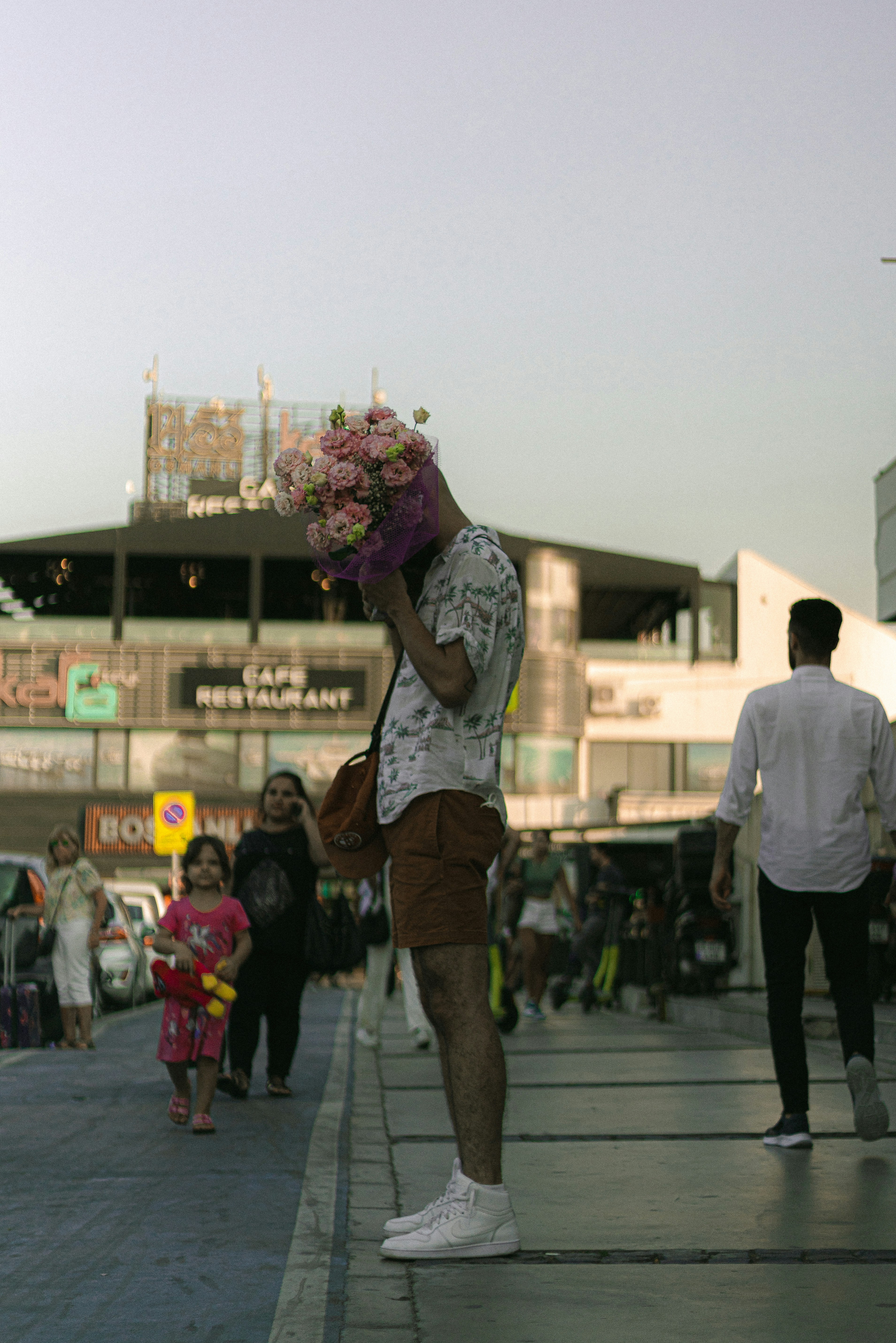 A man holding a large bouquet of flowers stands in a bustling urban environment, surrounded by pedestrians and shops. The scene captures the vibrancy of city life.