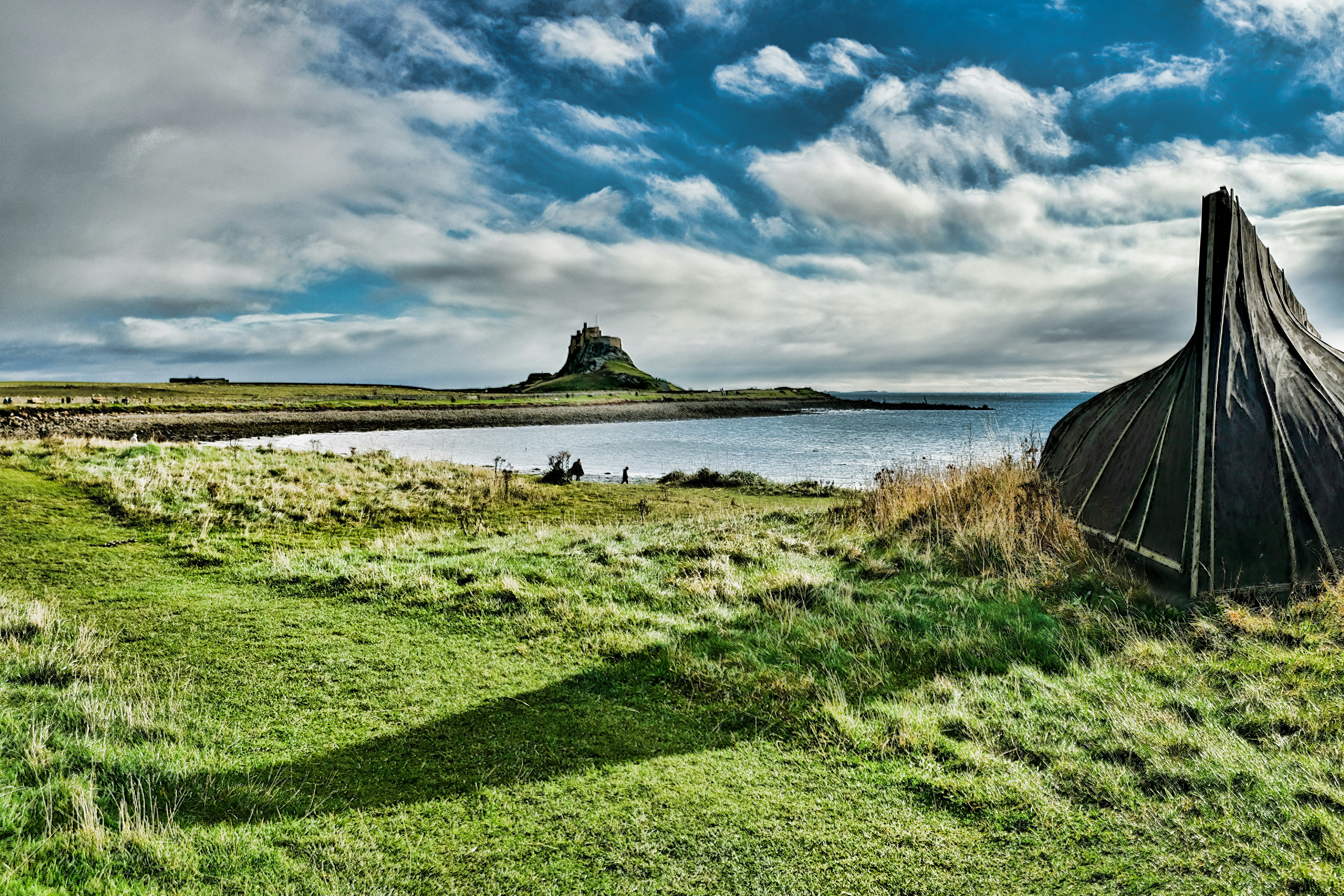 A weathered boat hull rests on grassy shores, overlooking a tranquil bay and a distant castle atop a hill.