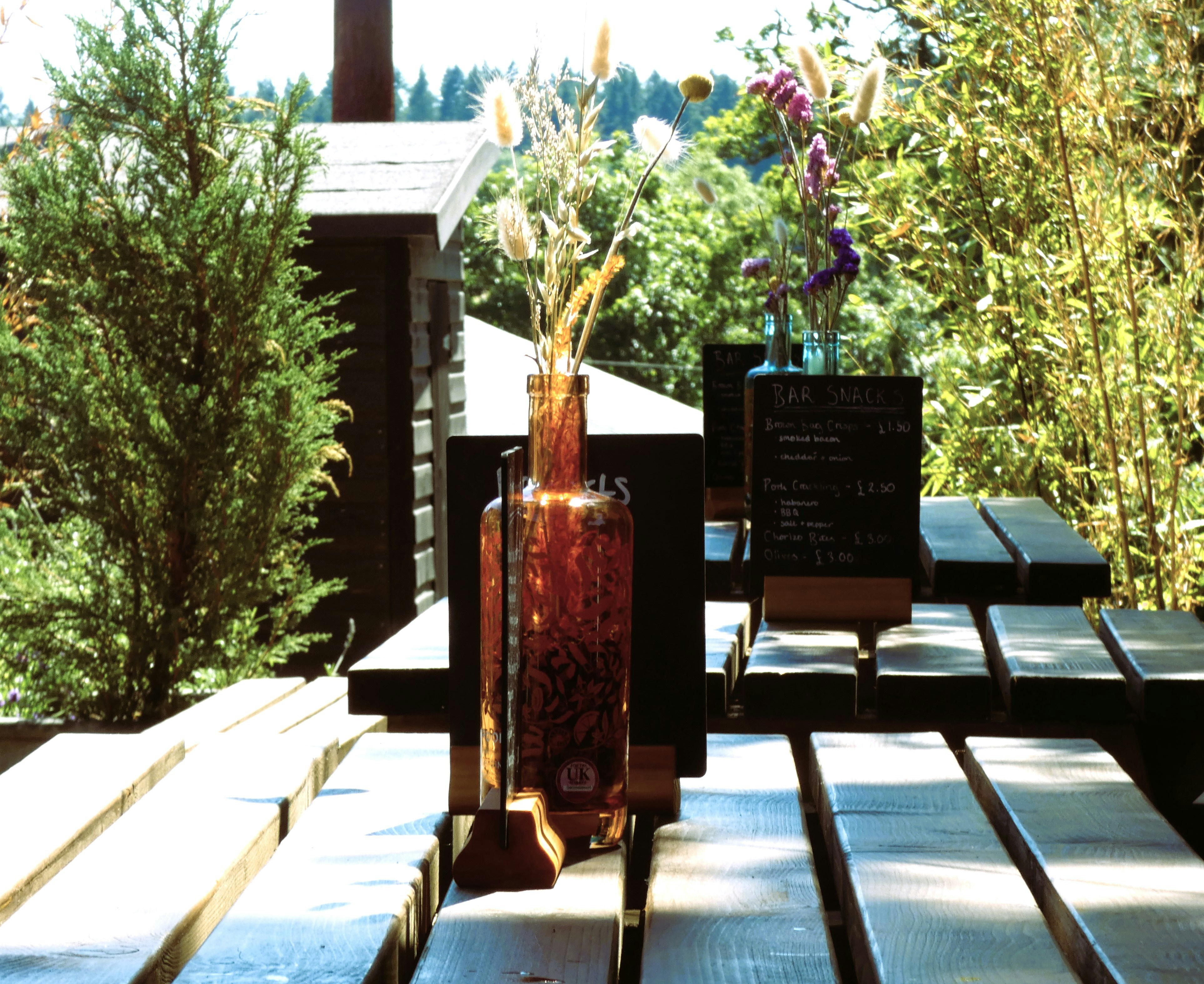 Glass vase with wildflowers on a wooden table under dappled sunlight, surrounded by lush greenery.
