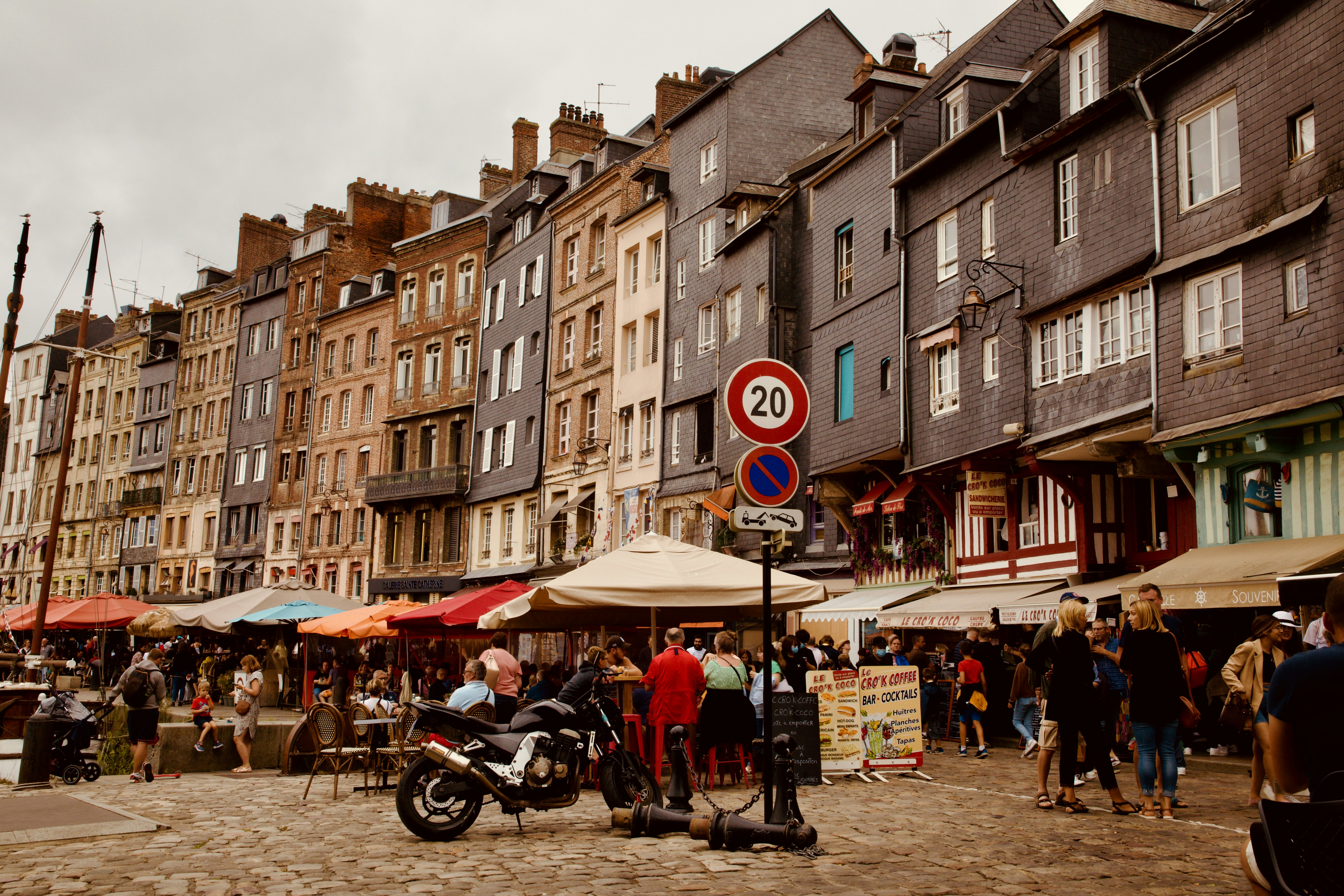 Colorful buildings line a bustling street in Honfleur, showcasing outdoor cafes and lively pedestrians. A motorcycle and traffic sign add to the vibrant urban scene.