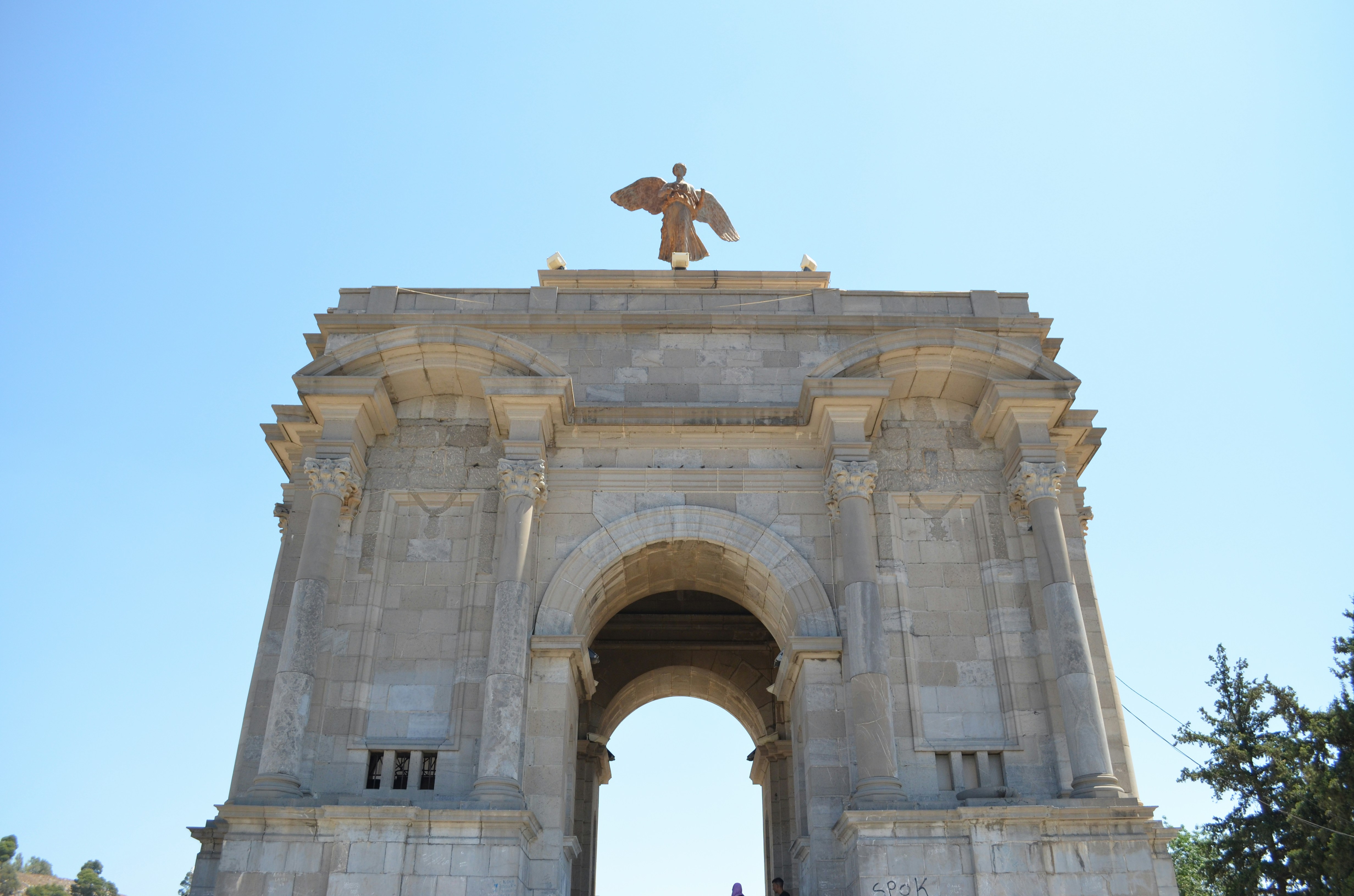 Stone arch monument crowned with a statue against a bright blue sky.