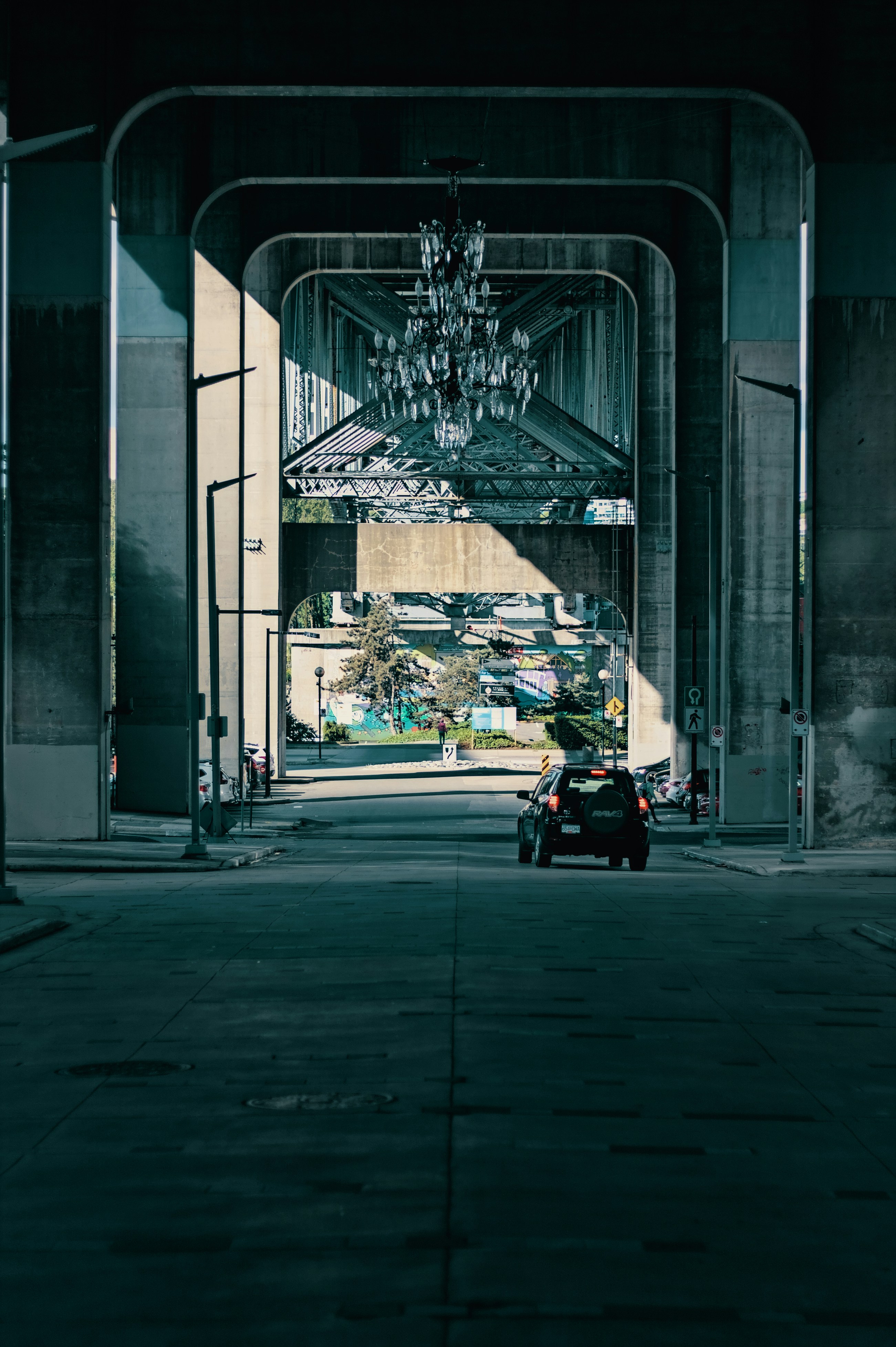 A sleek car drives through a shadowy underpass adorned with an unexpected chandelier, showcasing a blend of industrial architecture and artistic flair.