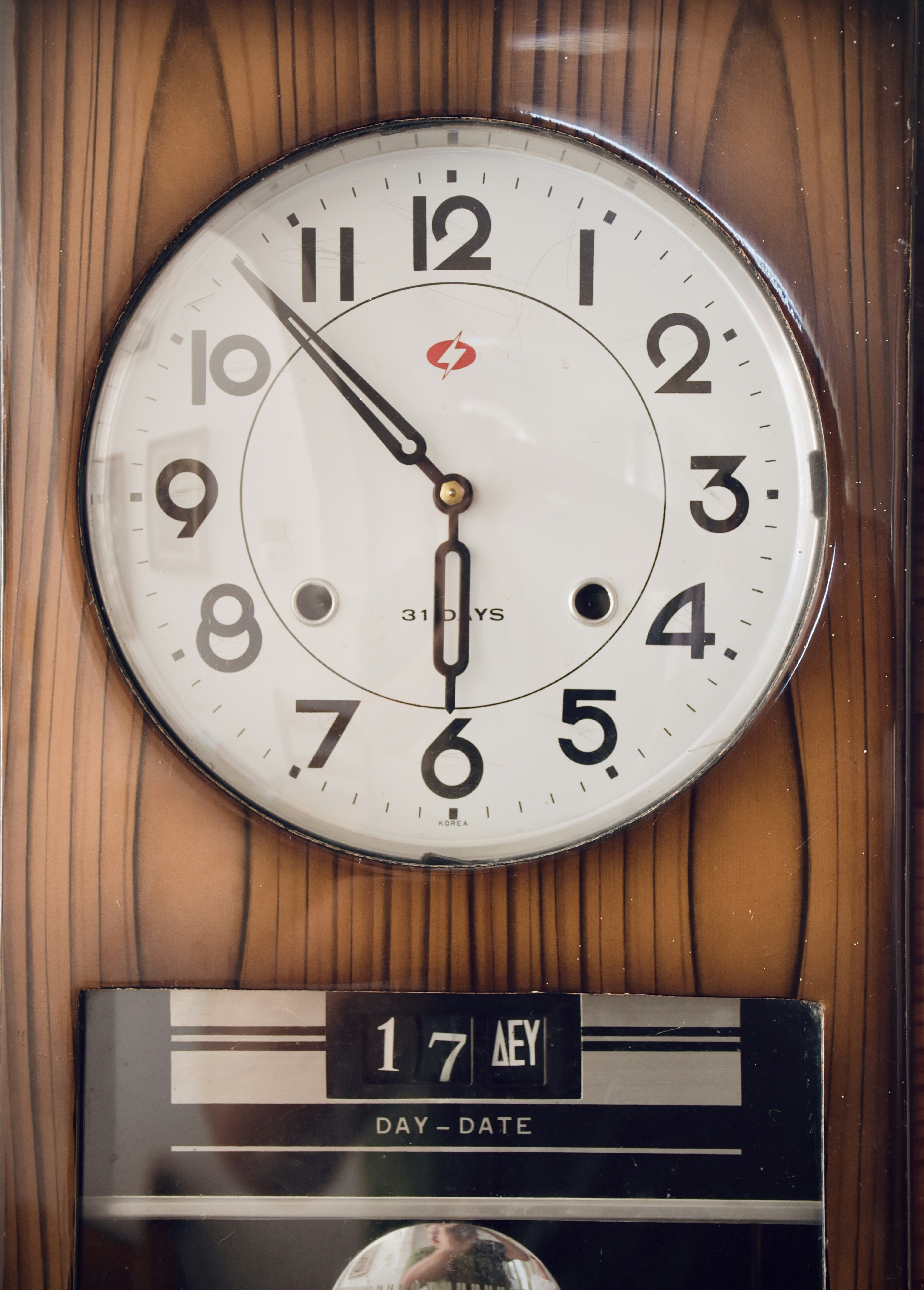 Classic wall clock featuring a minimalist design with a white face and black numerals, set against a wooden background. The clock displays the date along with the time.