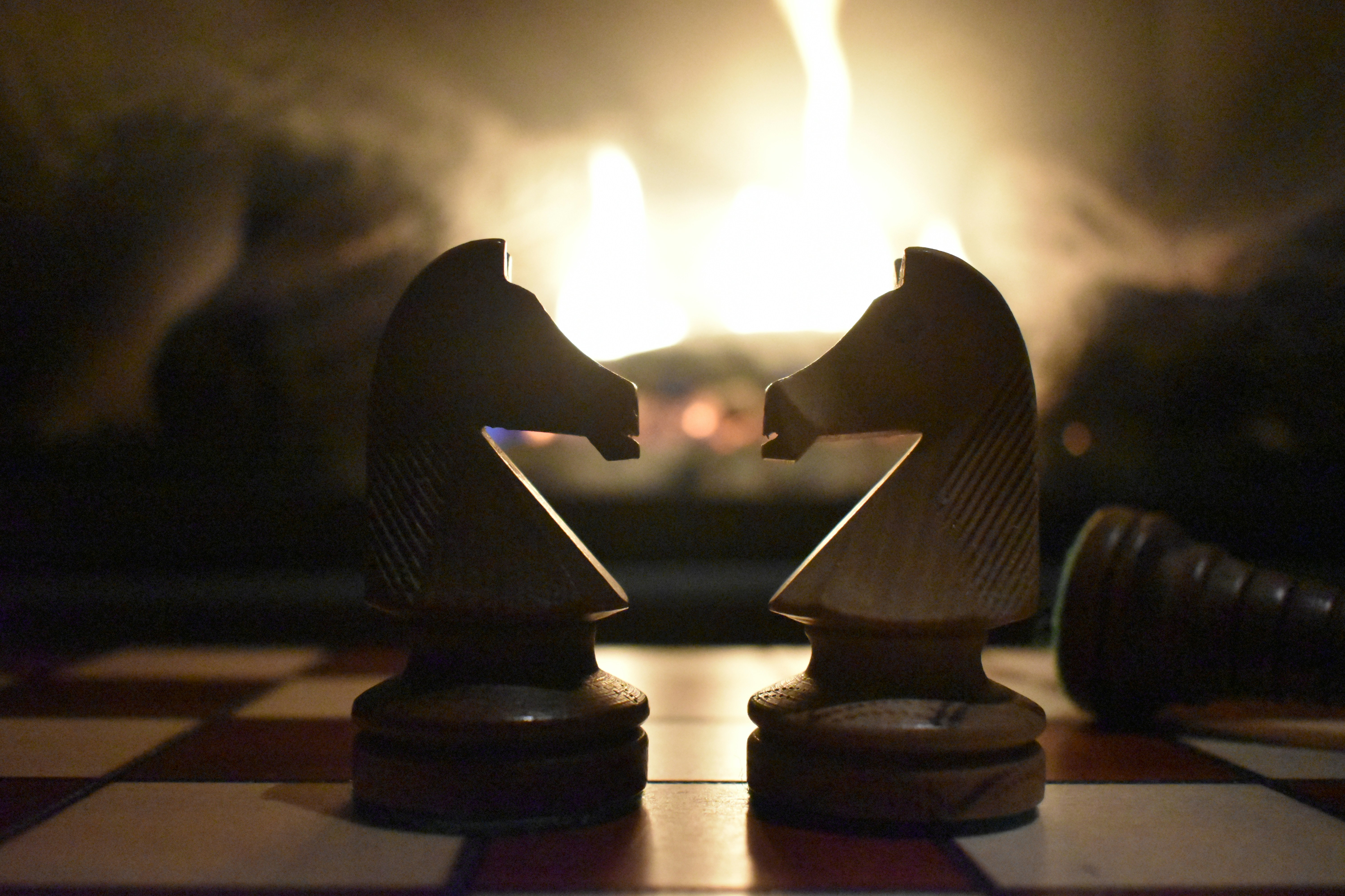 Silhouetted chess knights face each other with a bright fire in the background on a wooden chessboard.