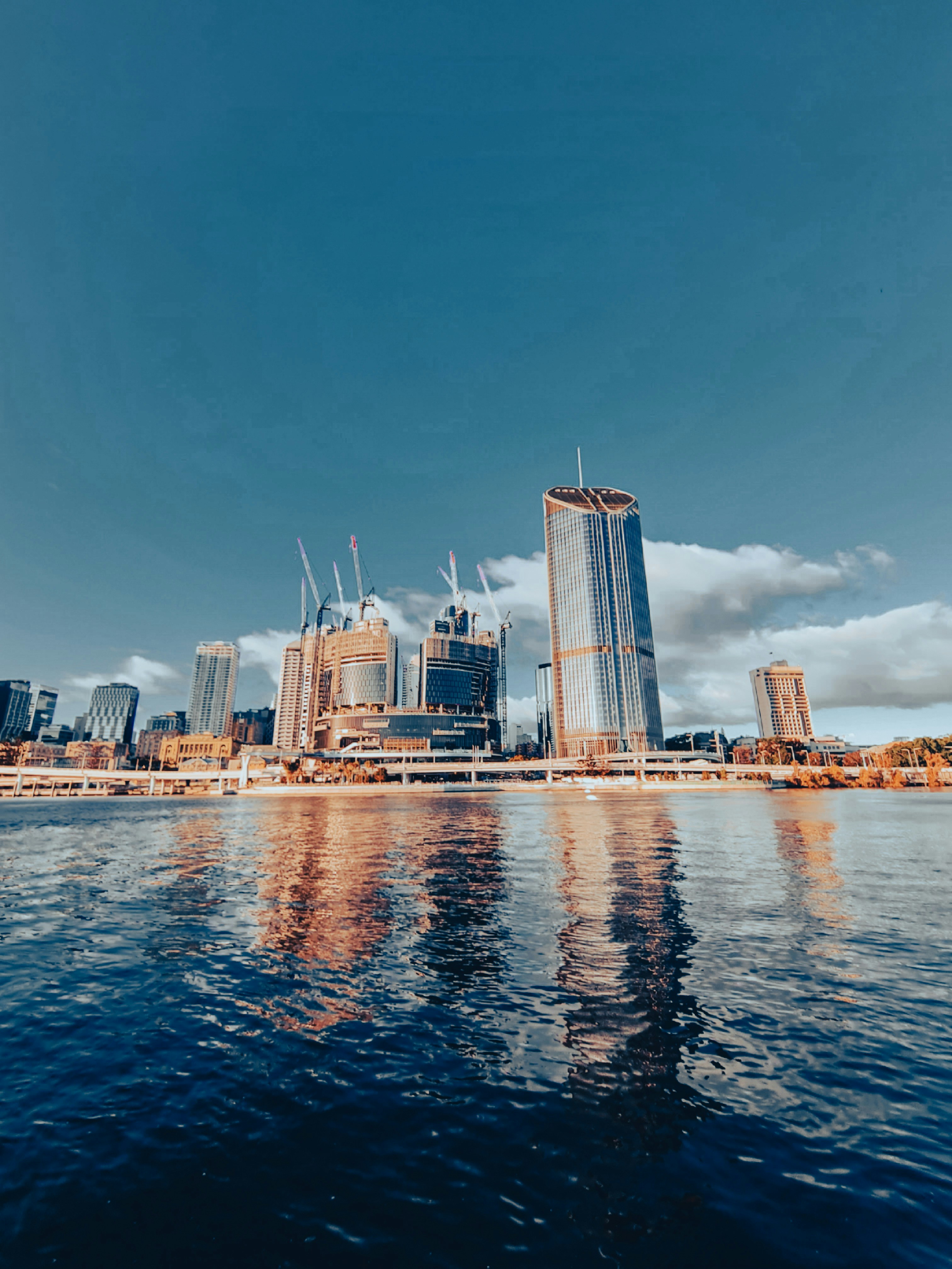 Modern skyline with construction cranes reflecting on calm water under a clear blue sky.