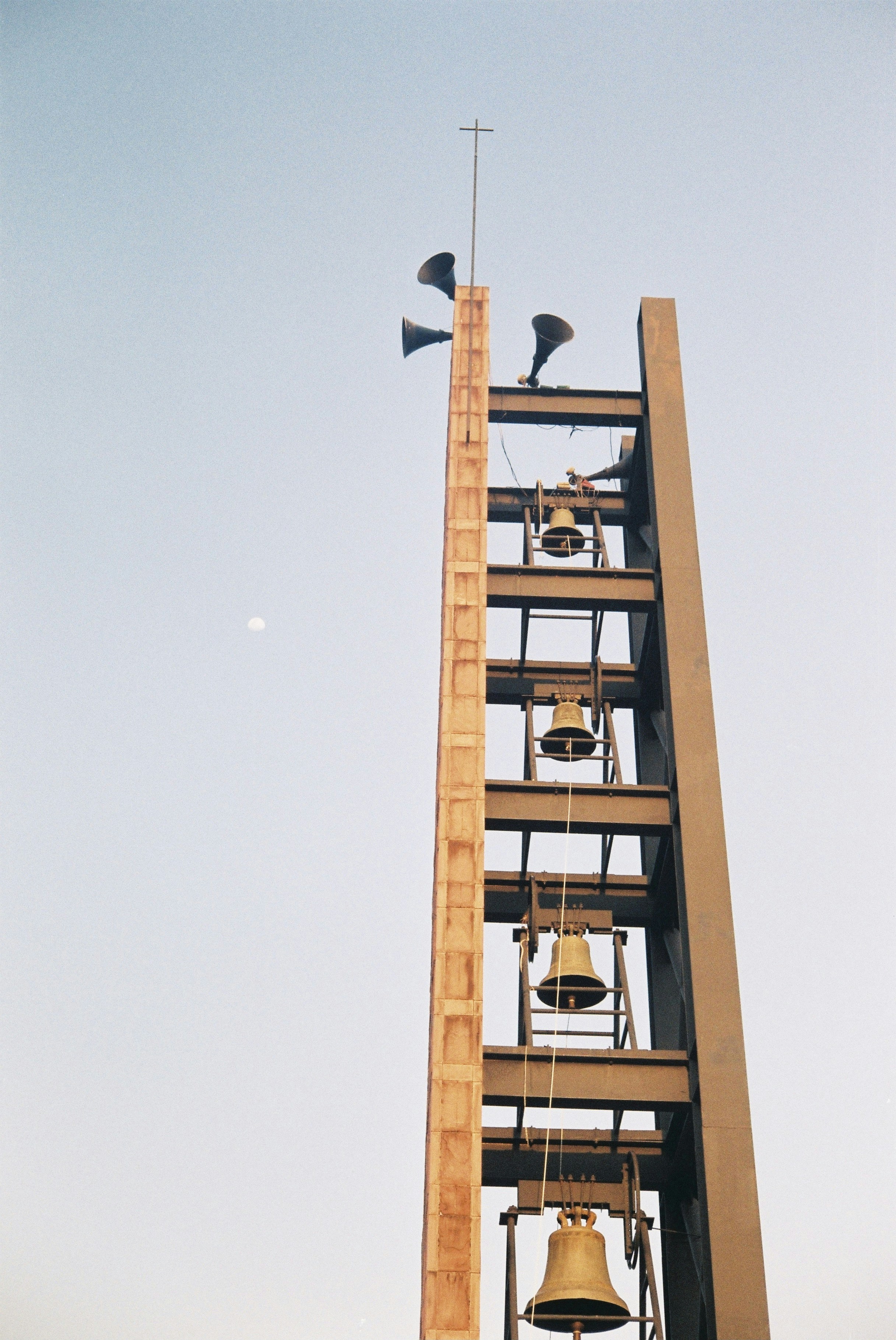 A towering bell structure adorned with multiple bells and speakers against a pale sky, capturing a moment of stillness. The moon subtly glimmers in the background.