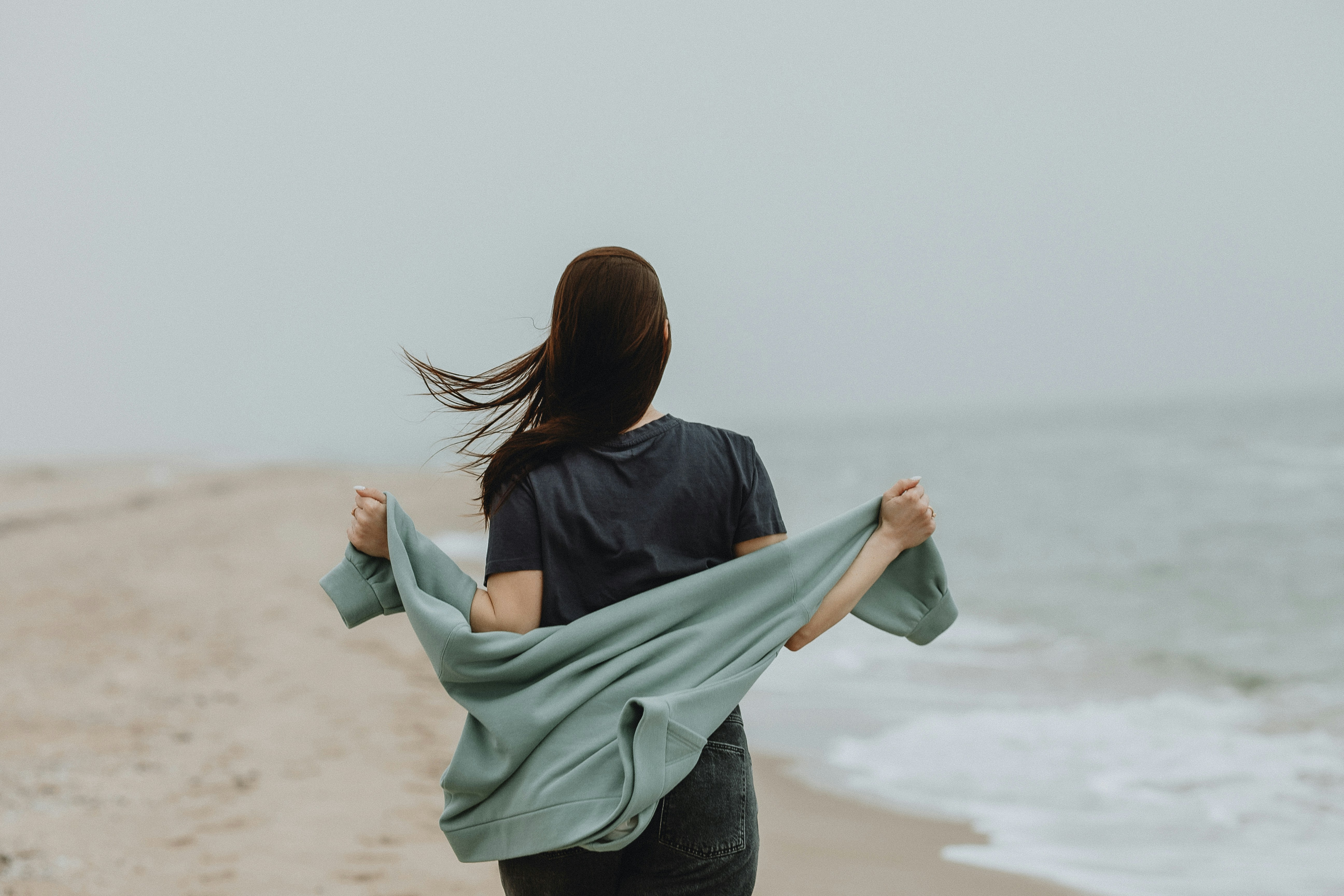Woman in a casual outfit stands on a sandy beach, holding a light sweater as her hair flows in the breeze, with gentle waves lapping at the shore.
