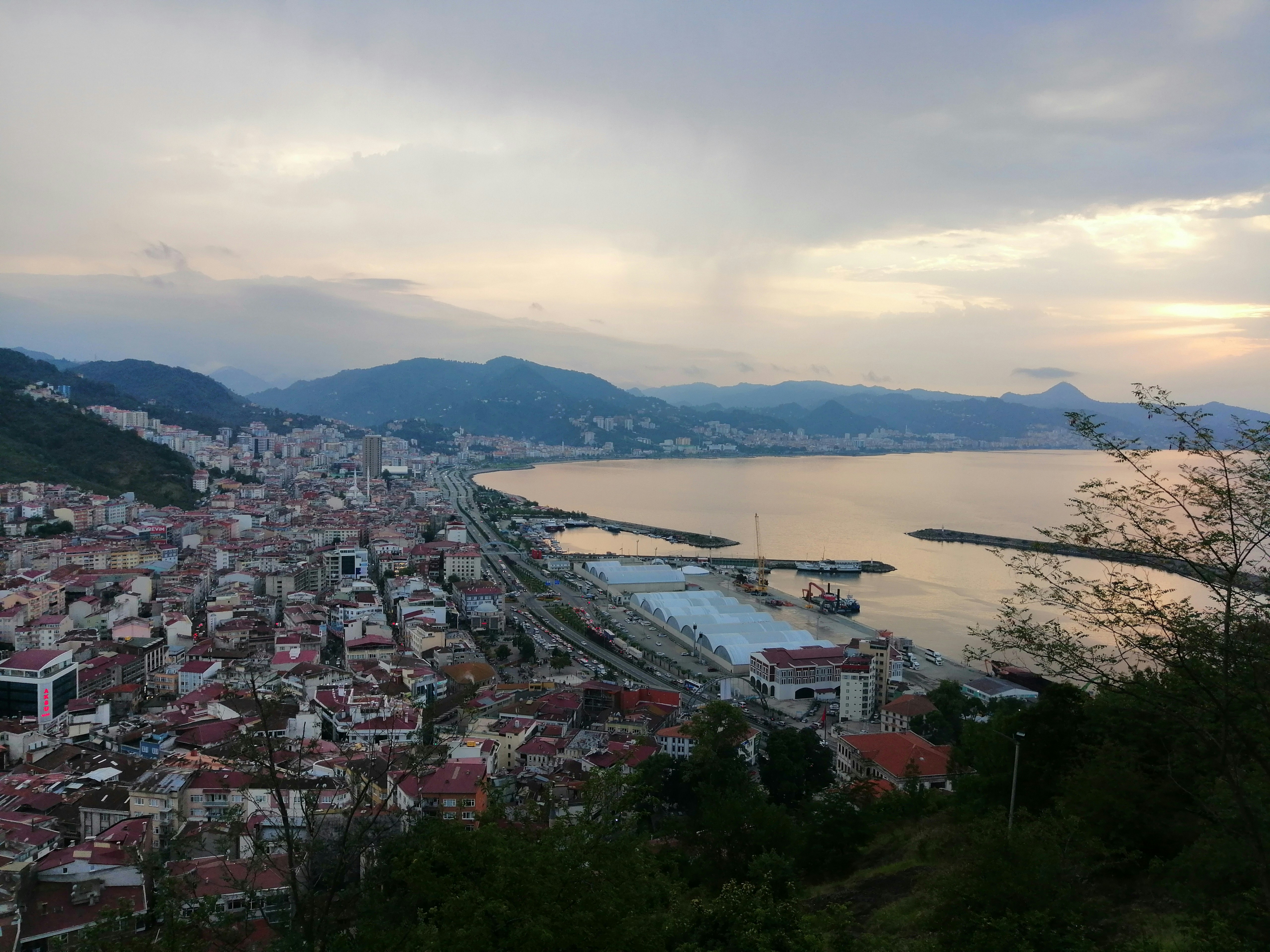 Panoramic view of a coastal city at dusk with mountains in the background and gentle waves lapping against a pier.