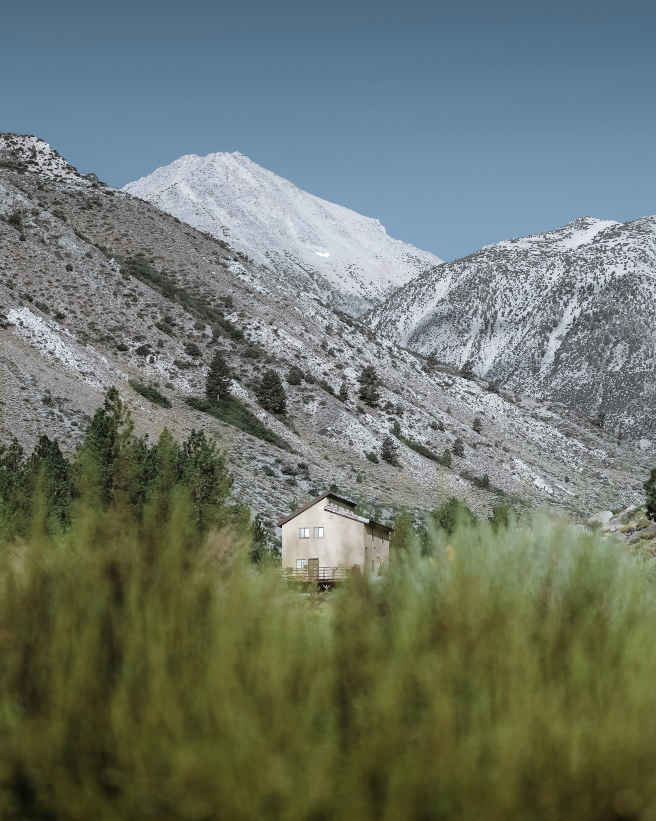 A rustic house nestled in a lush valley, framed by towering mountains and a clear blue sky. The scene conveys a sense of tranquility and harmony with nature.