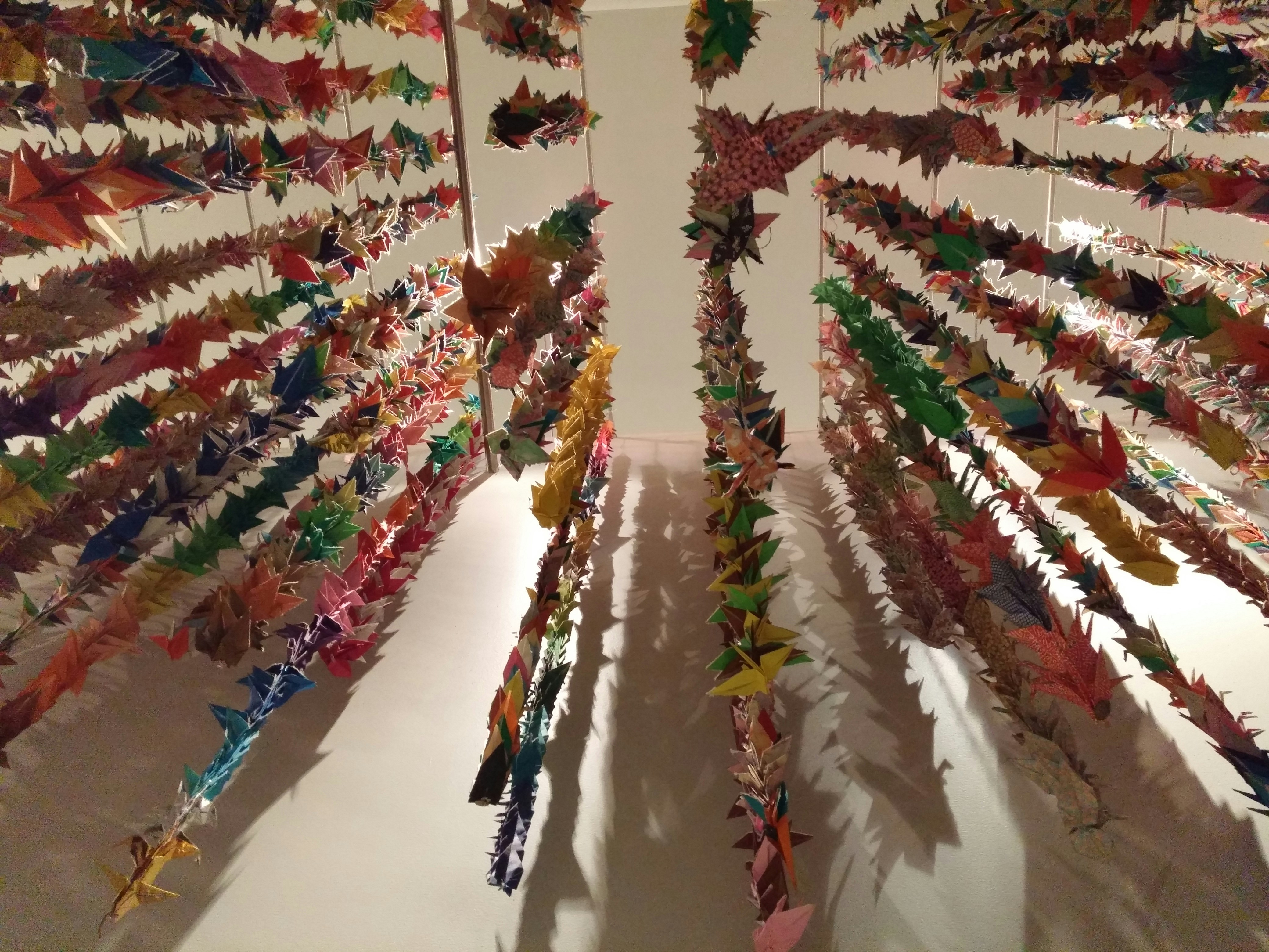 Colorful origami cranes suspended in mid-air create a vibrant installation, casting intricate shadows on the floor.