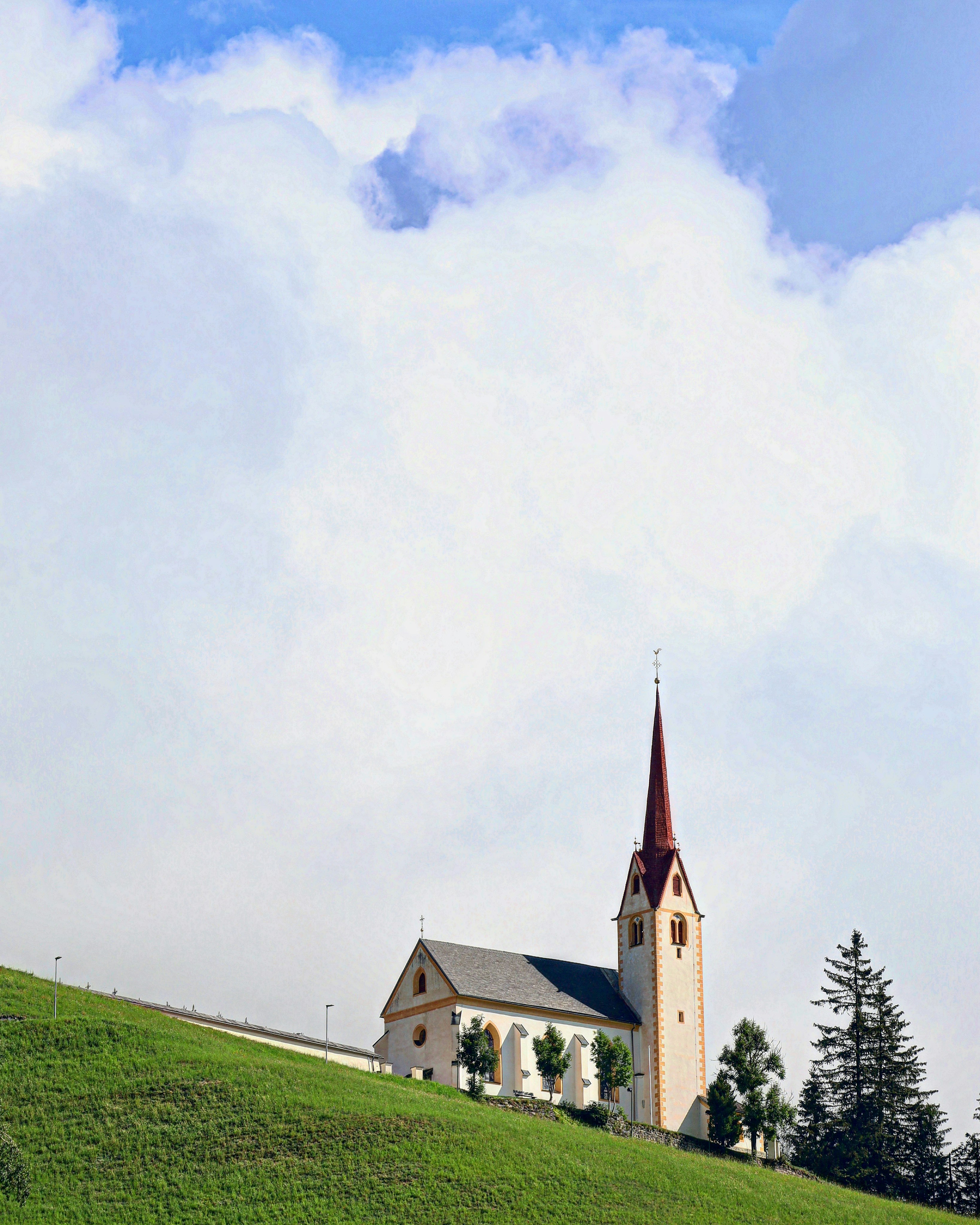 A quaint church with a tall steeple nestled on a green hillside, framed by a backdrop of soft clouds.
