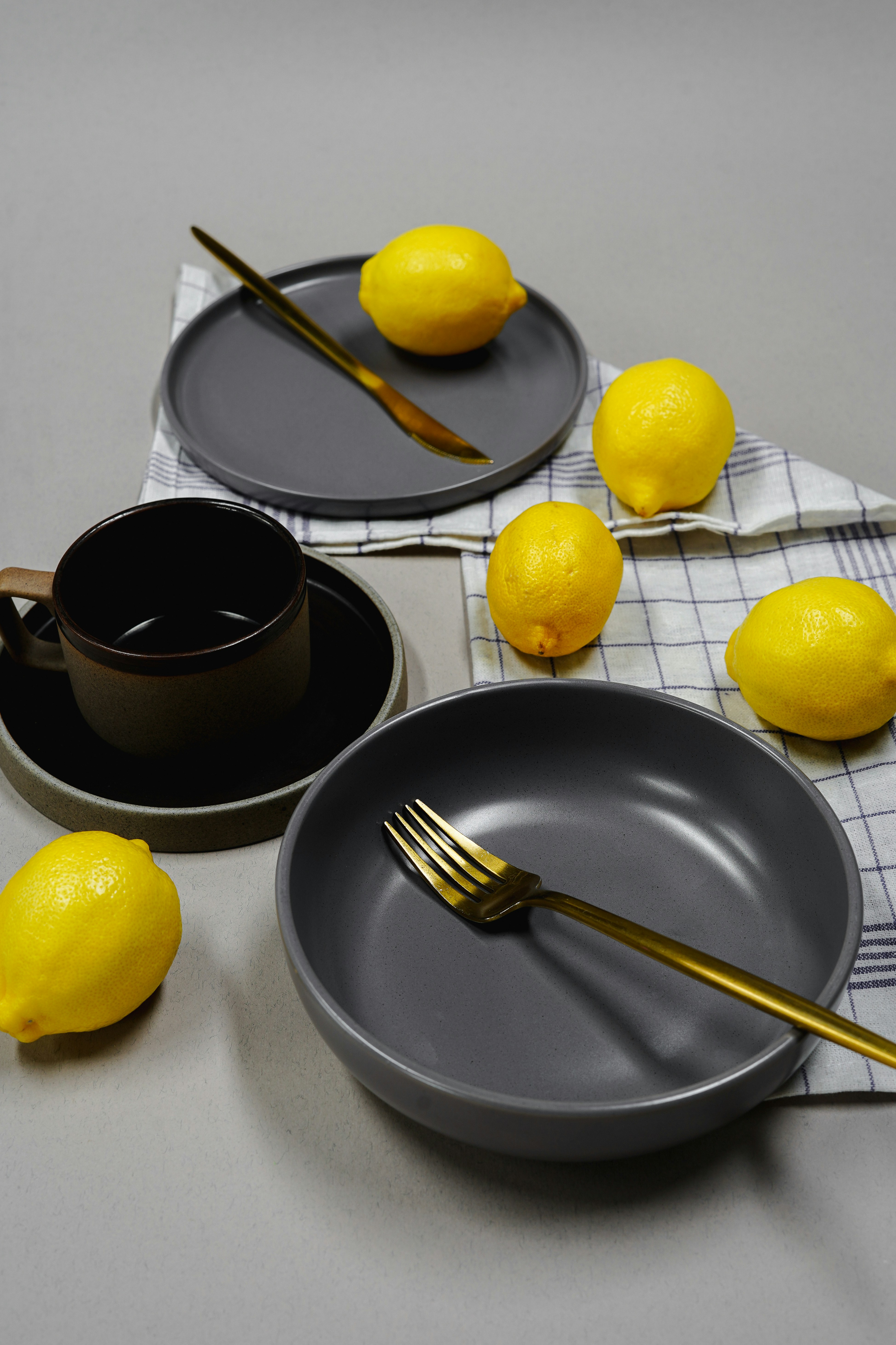 A minimalist arrangement featuring gray plates, a dark cup, and vibrant lemons on a checkered cloth. This setup highlights the contrast between the earthy tones and the bright yellow fruit.