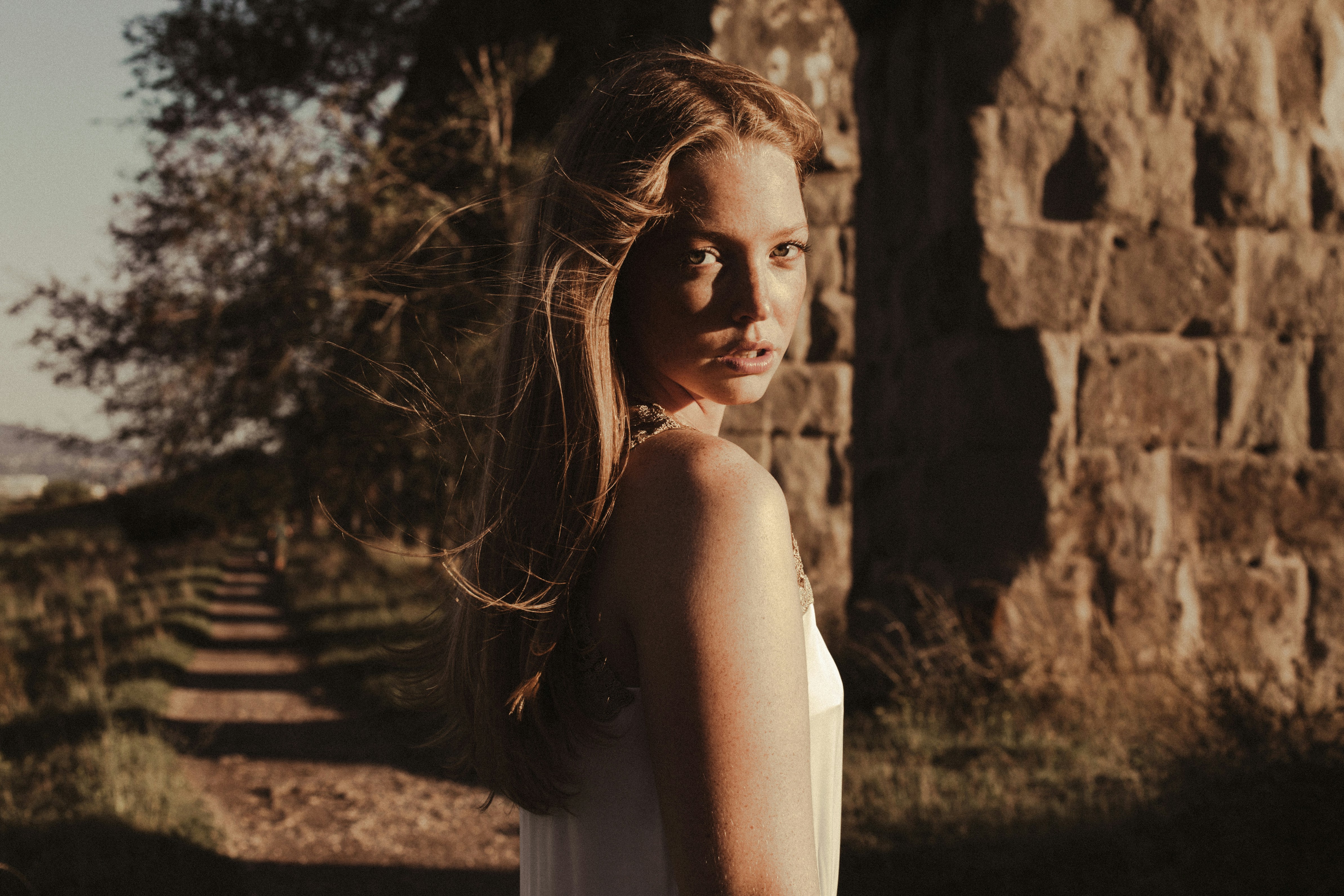 Woman in a white dress stands in warm, late afternoon sunlight with a stone wall behind her.