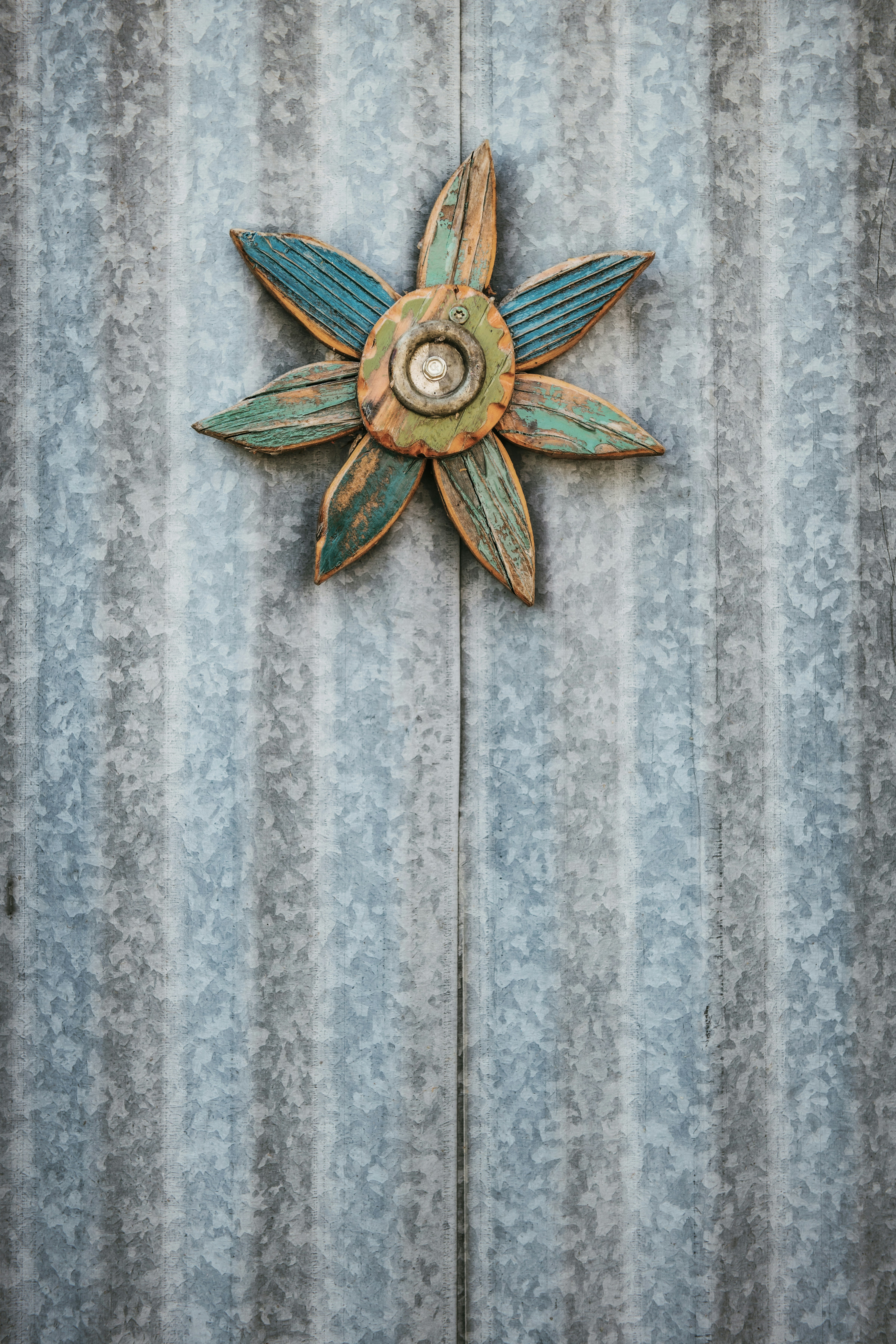A decorative metal flower with blue and orange accents mounted on a textured, corrugated metal background.