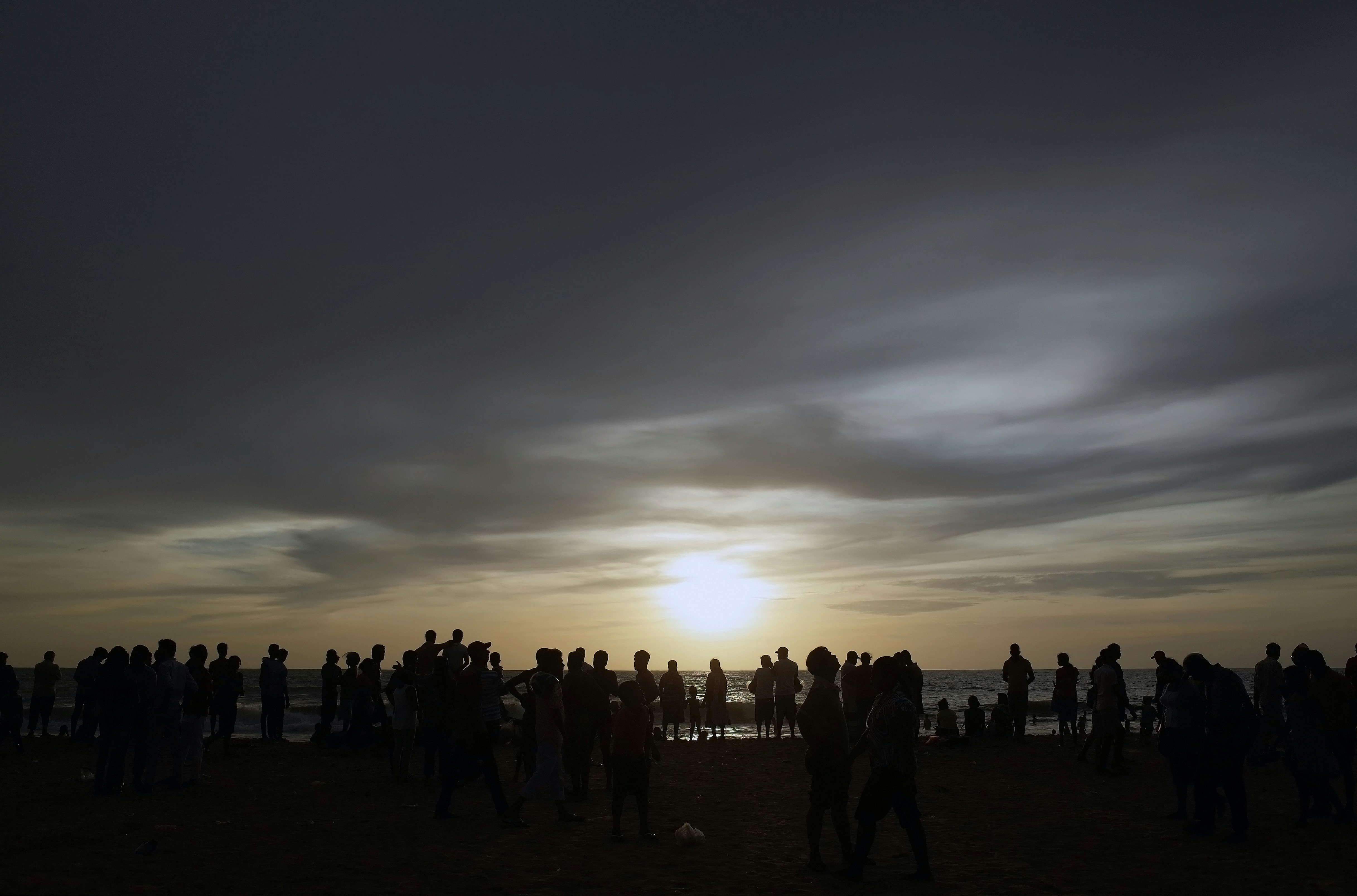 Silhouetted beachgoers line the shore as a low sun sinks toward the horizon, painting the sky with muted gold and sweeping clouds.