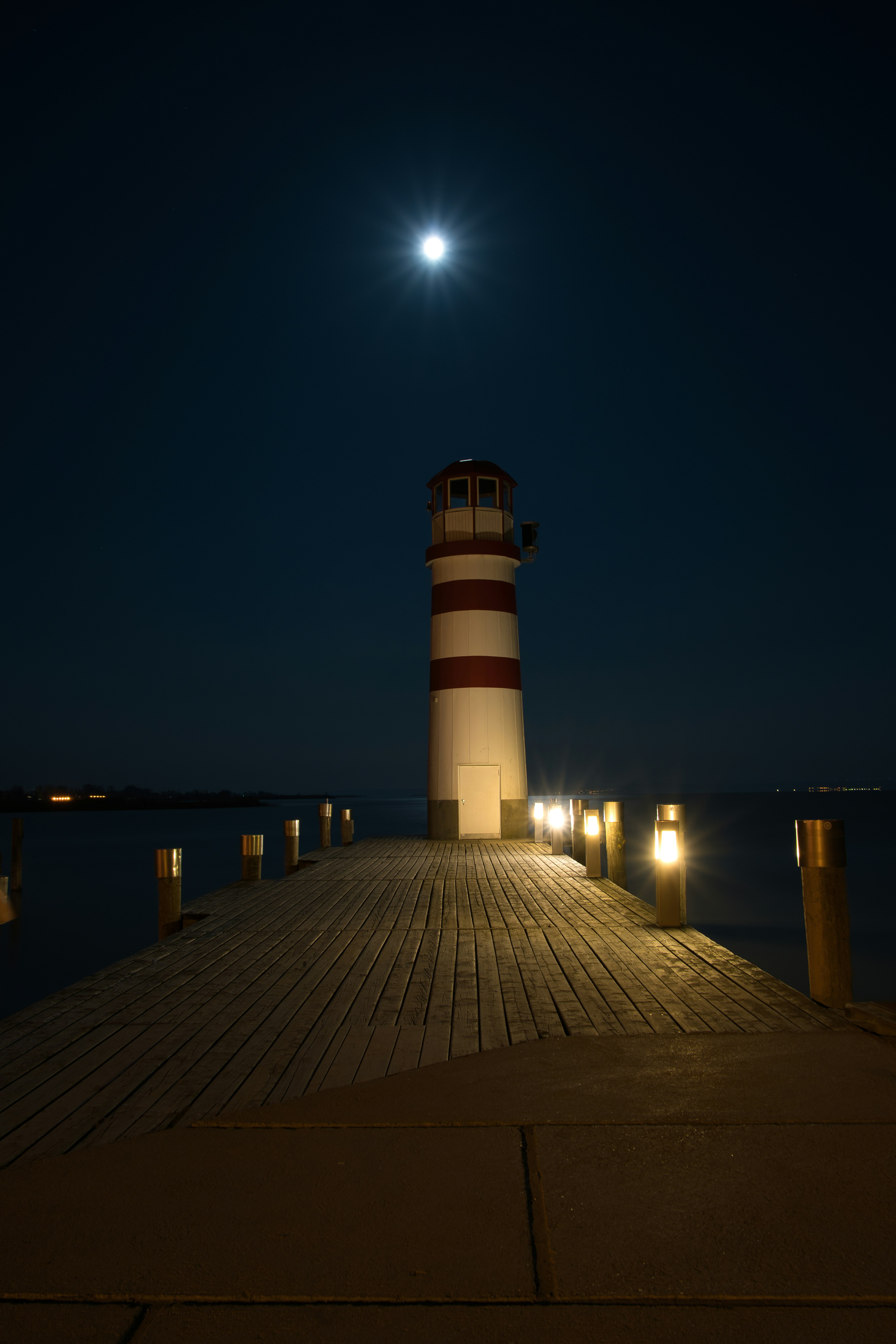 A lighthouse stands tall on a wooden pier illuminated by moonlight, with gentle reflections on the water's surface.